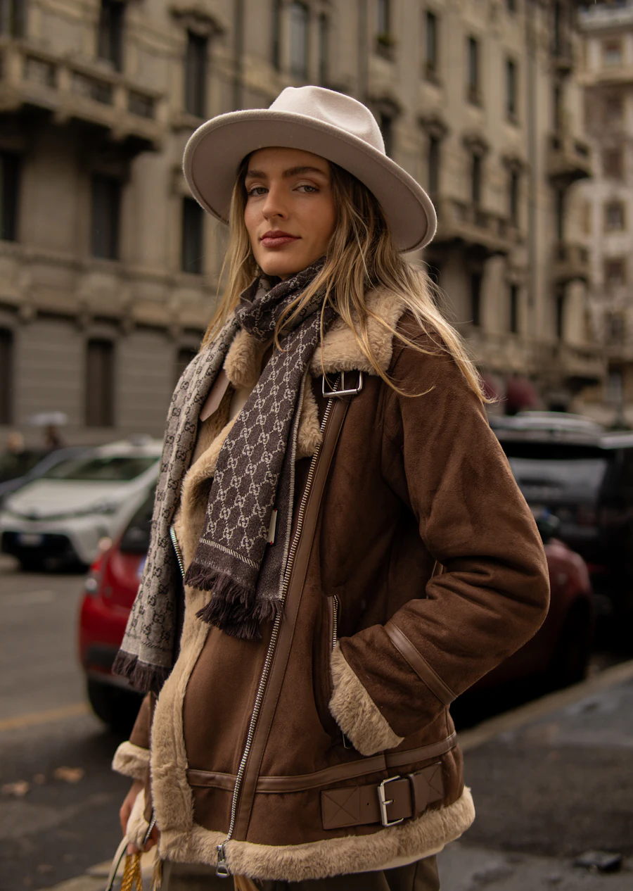 Fashion portrait of a woman in a wide-brim hat, brown shearling coat, and patterned scarf standing on a city street. Soft-focus buildings and parked cars frame the scene behind her.