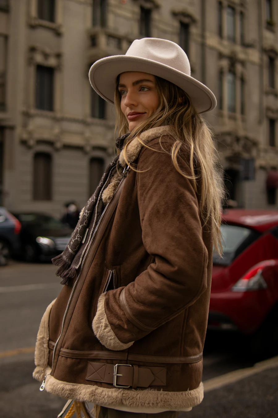 A woman in a brown shearling-lined coat and light wide-brim hat stands on a city street, looking over her shoulder. Soft-focus buildings and parked cars form the background.