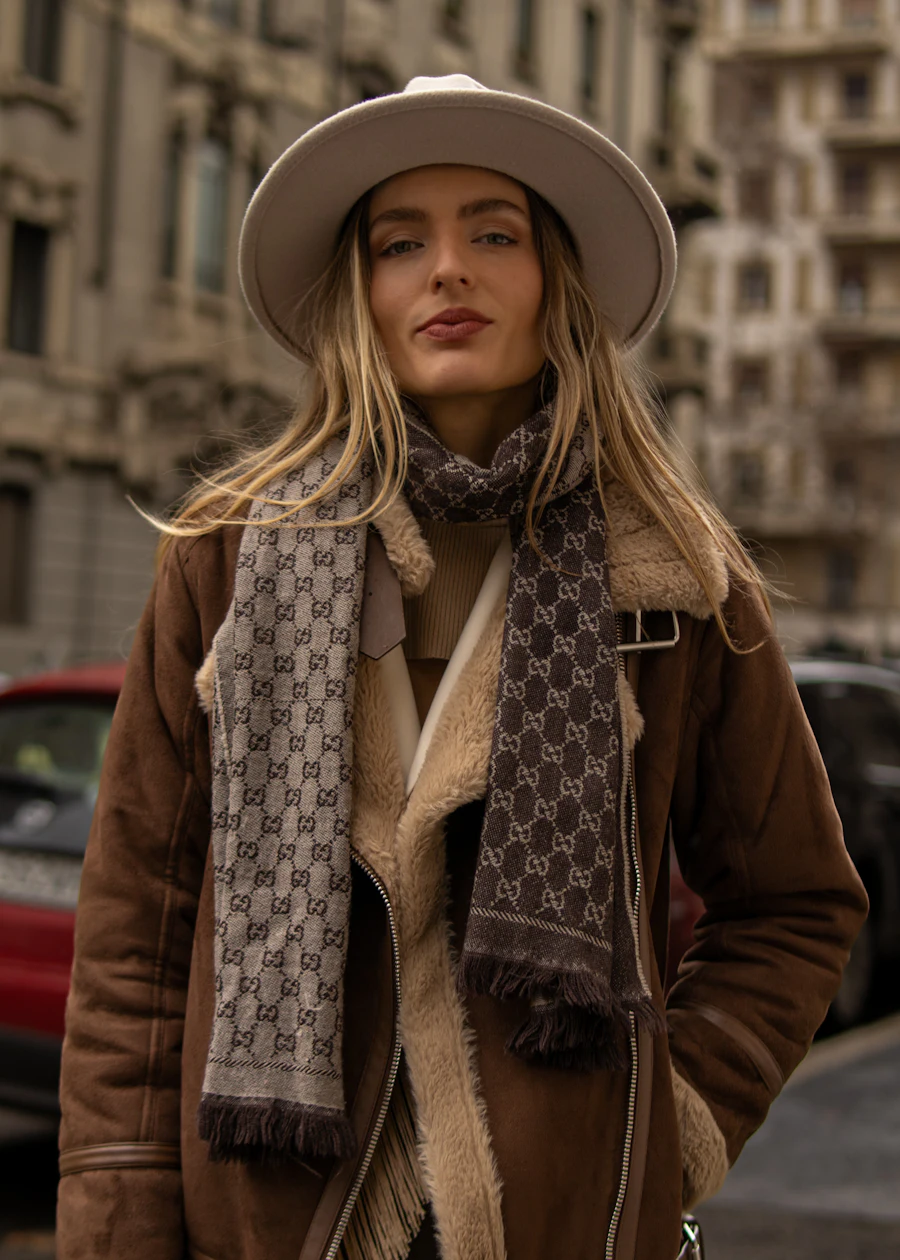 A woman in a wide-brim hat and layered brown coat stands on a city street, wearing patterned scarves. Softly blurred buildings and a red car form the background.