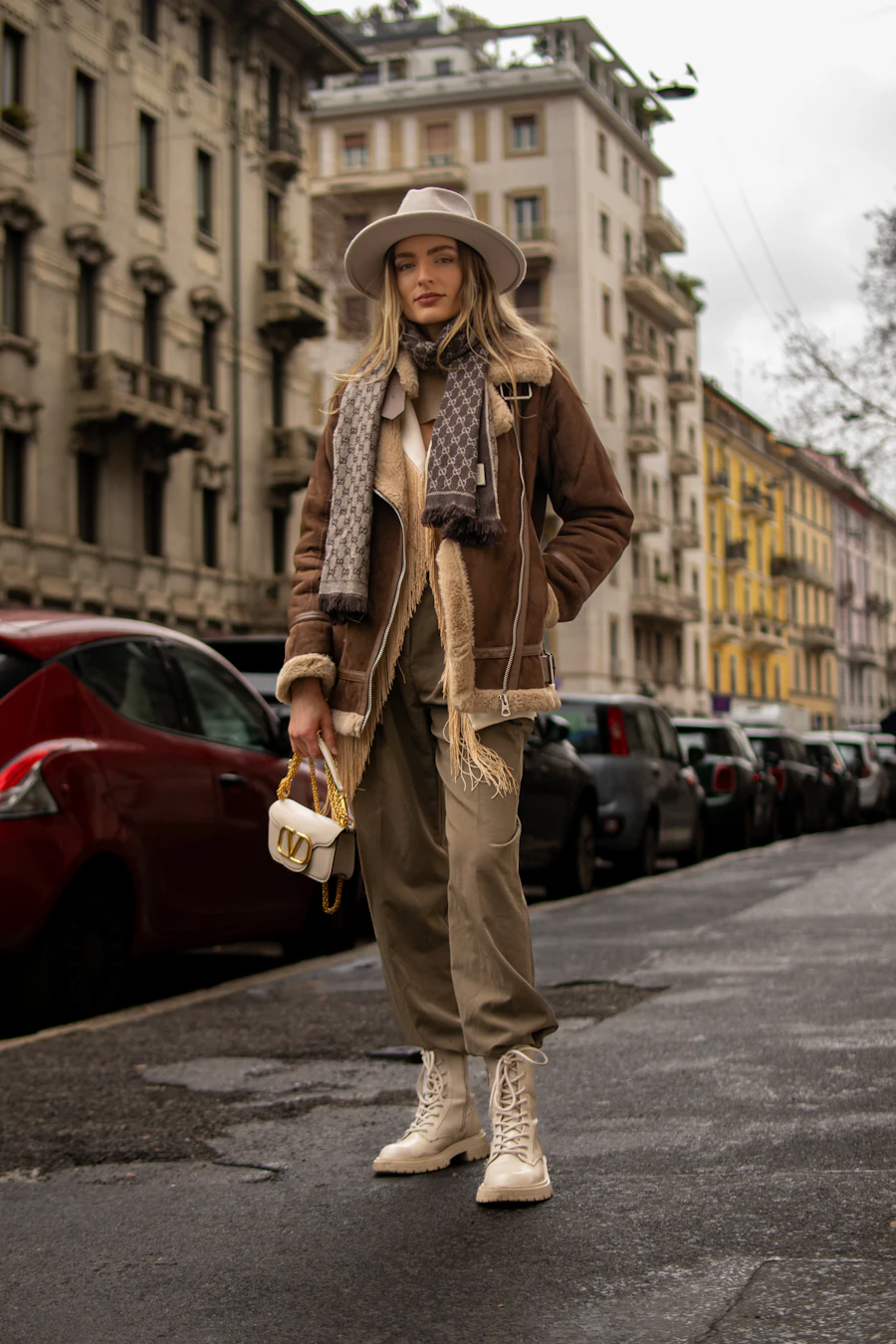 A woman in a wide-brim hat and shearling jacket stands on a wet city street with parked cars and old apartment buildings behind her. She holds a small white handbag and wears lace-up boots.