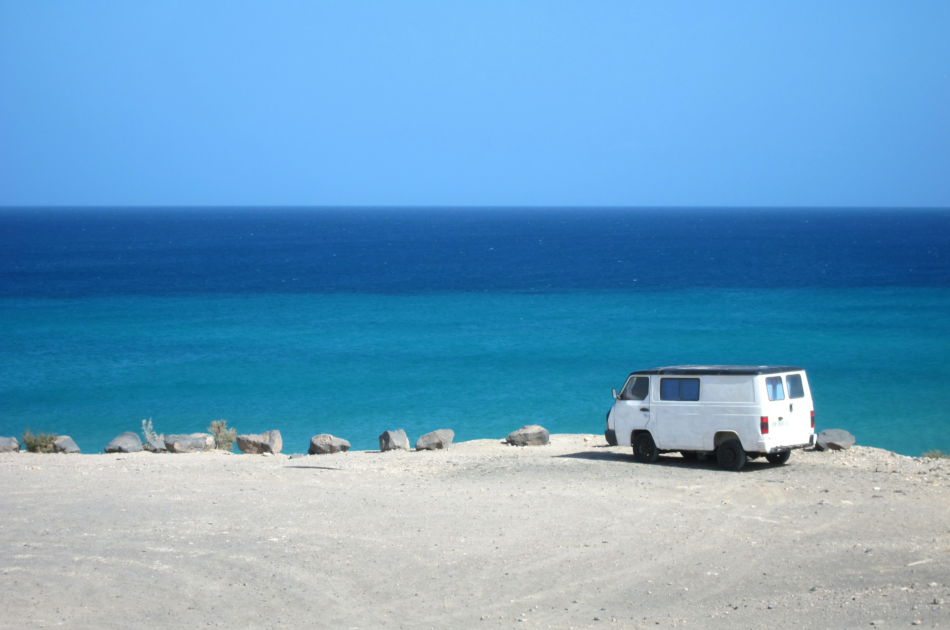 A white van sits on a sandy beach with turquoise water and a clear blue sky.