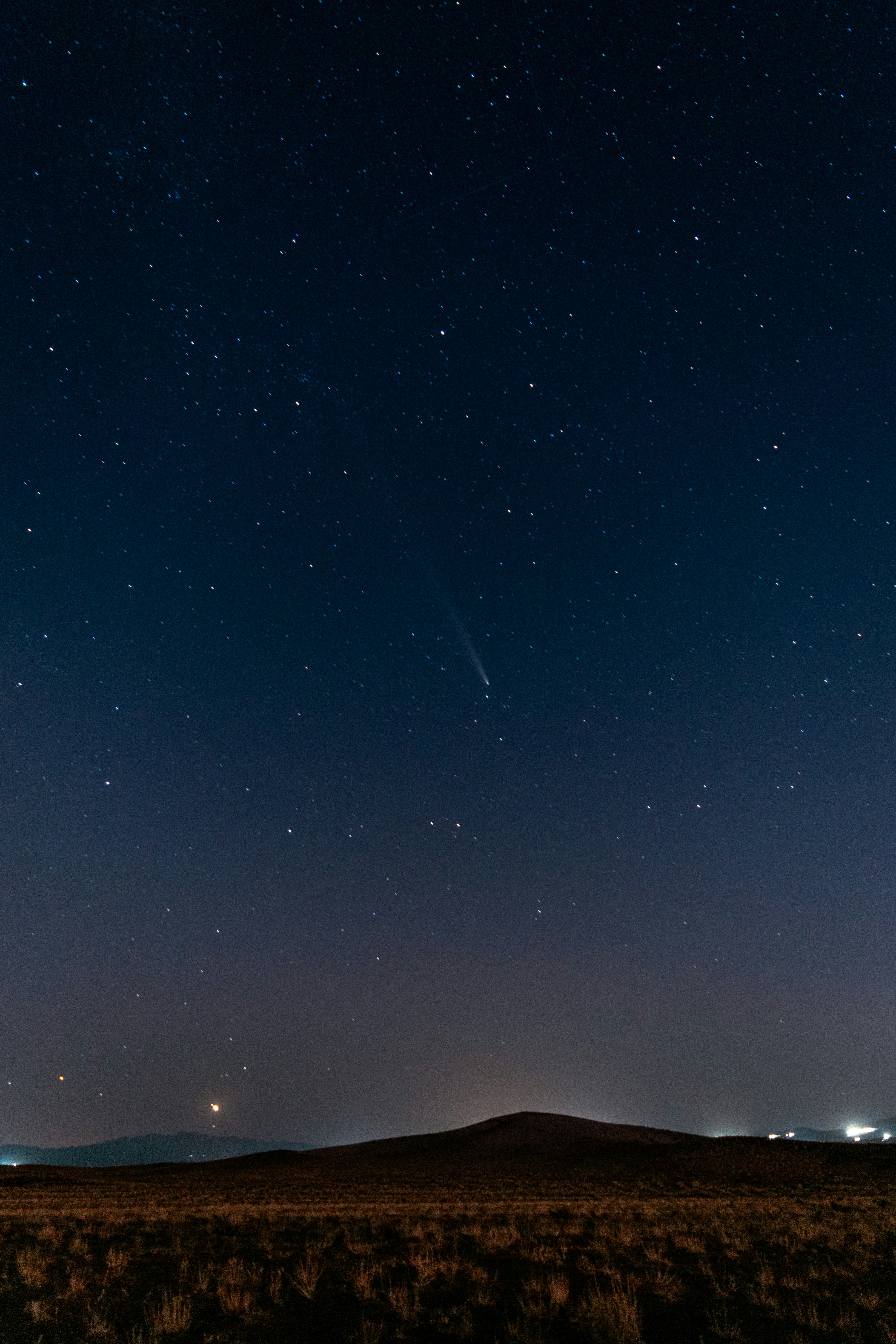 A comet streaks across the starry night sky above a vast, open landscape, illuminating the horizon with its faint glow.