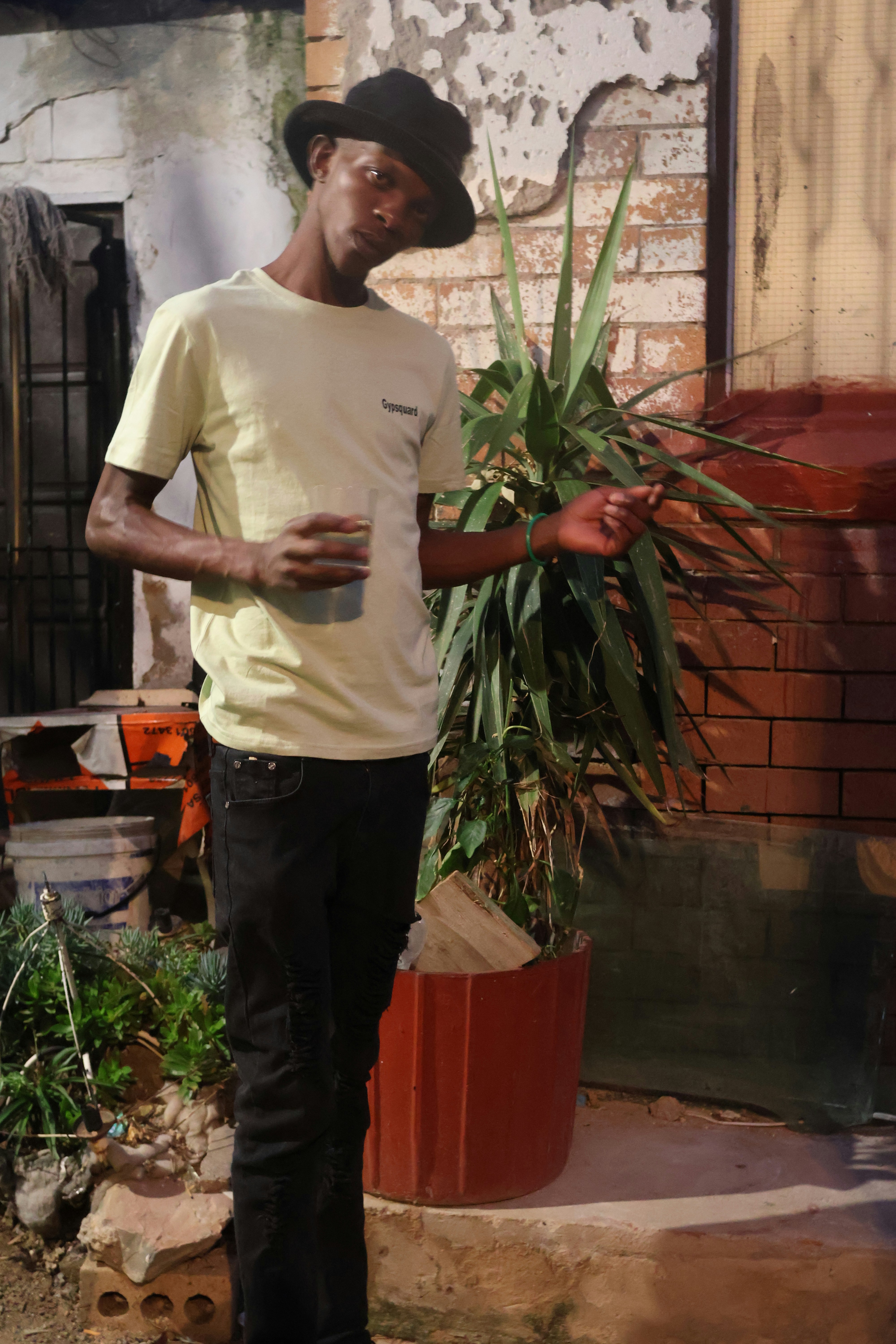 Sunlit Portrait by Brick and Plant: A Man in Pale Tee and Fedora