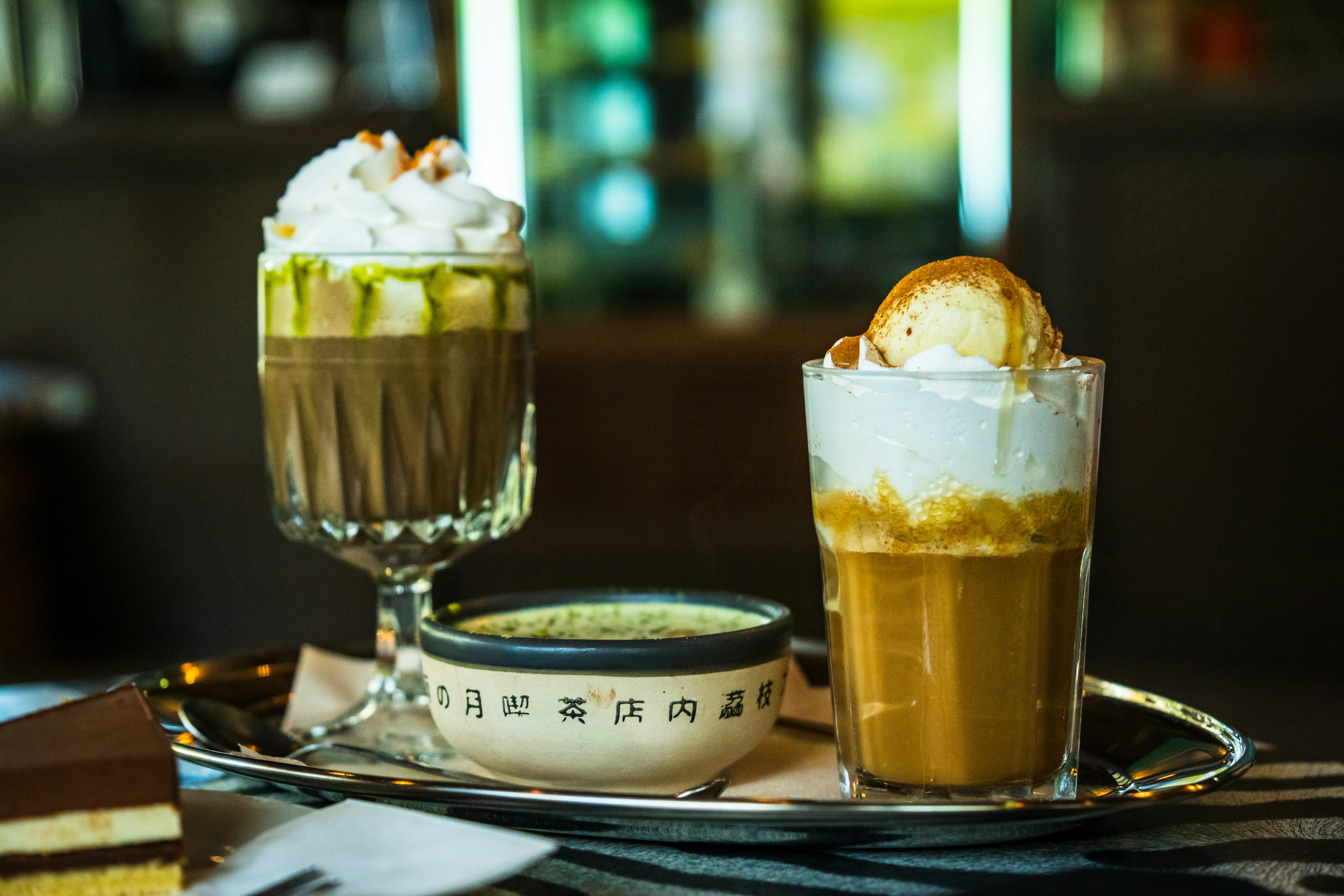 Two dessert beverages sit on a reflective tray: a pistachio-topped frappe with whipped cream and a caramel latte crowned with foam, accompanied by a small cup and a slice of cake.