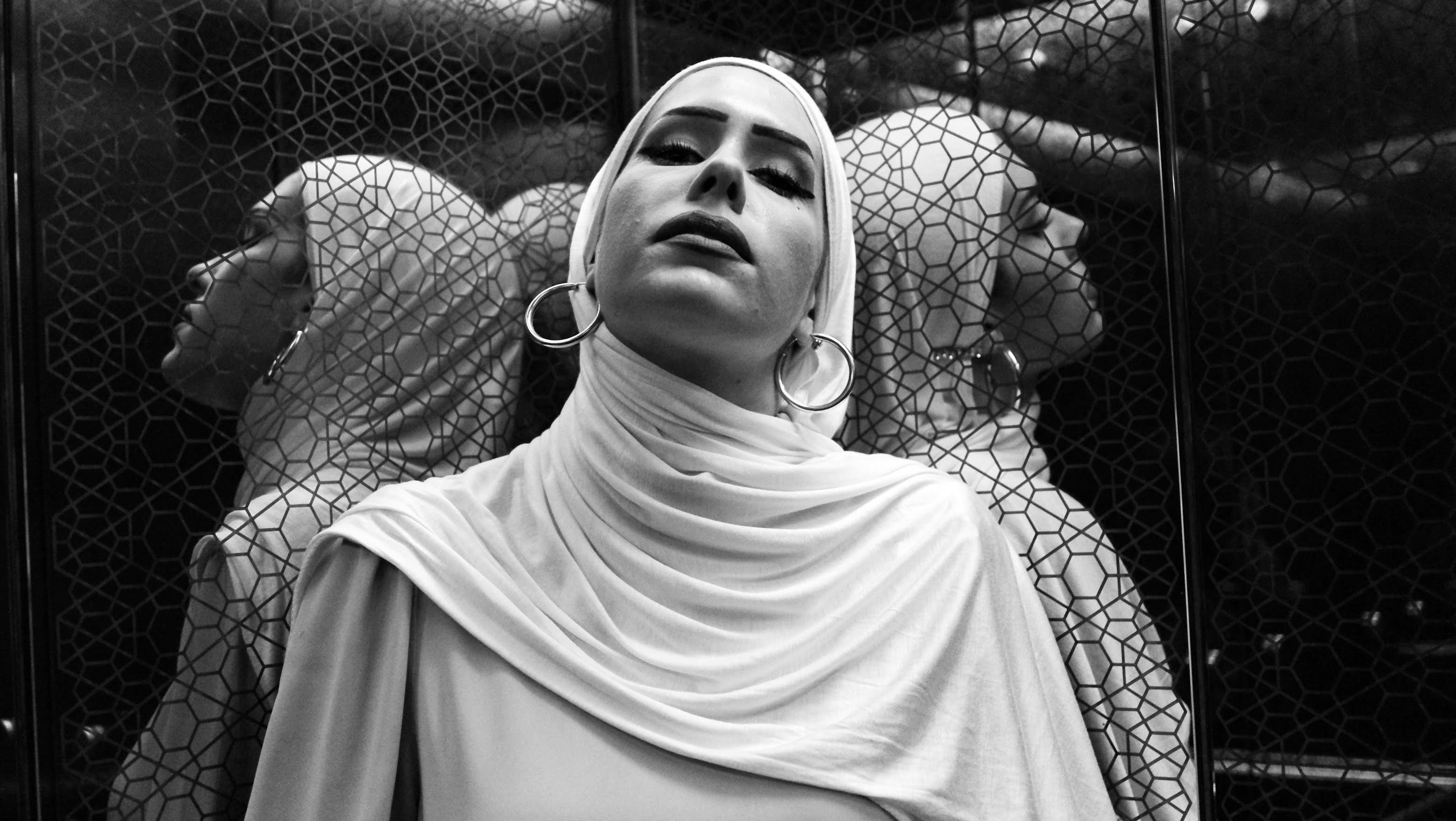 Woman in a white hijab stands between mirrored walls, creating multiple reflections under soft lighting.