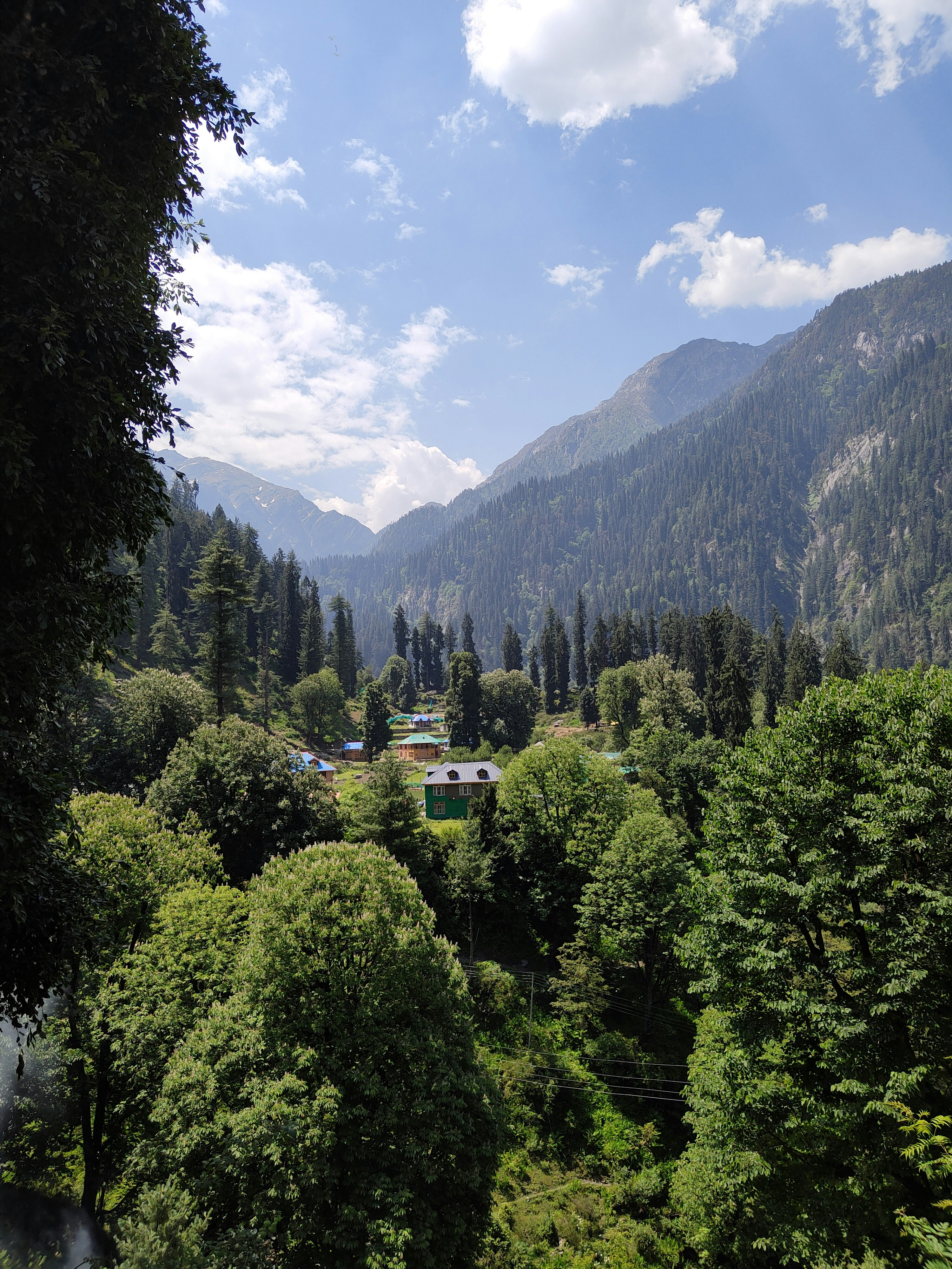 Sunlit green valley with dense forest and distant mountains under a partly cloudy sky.