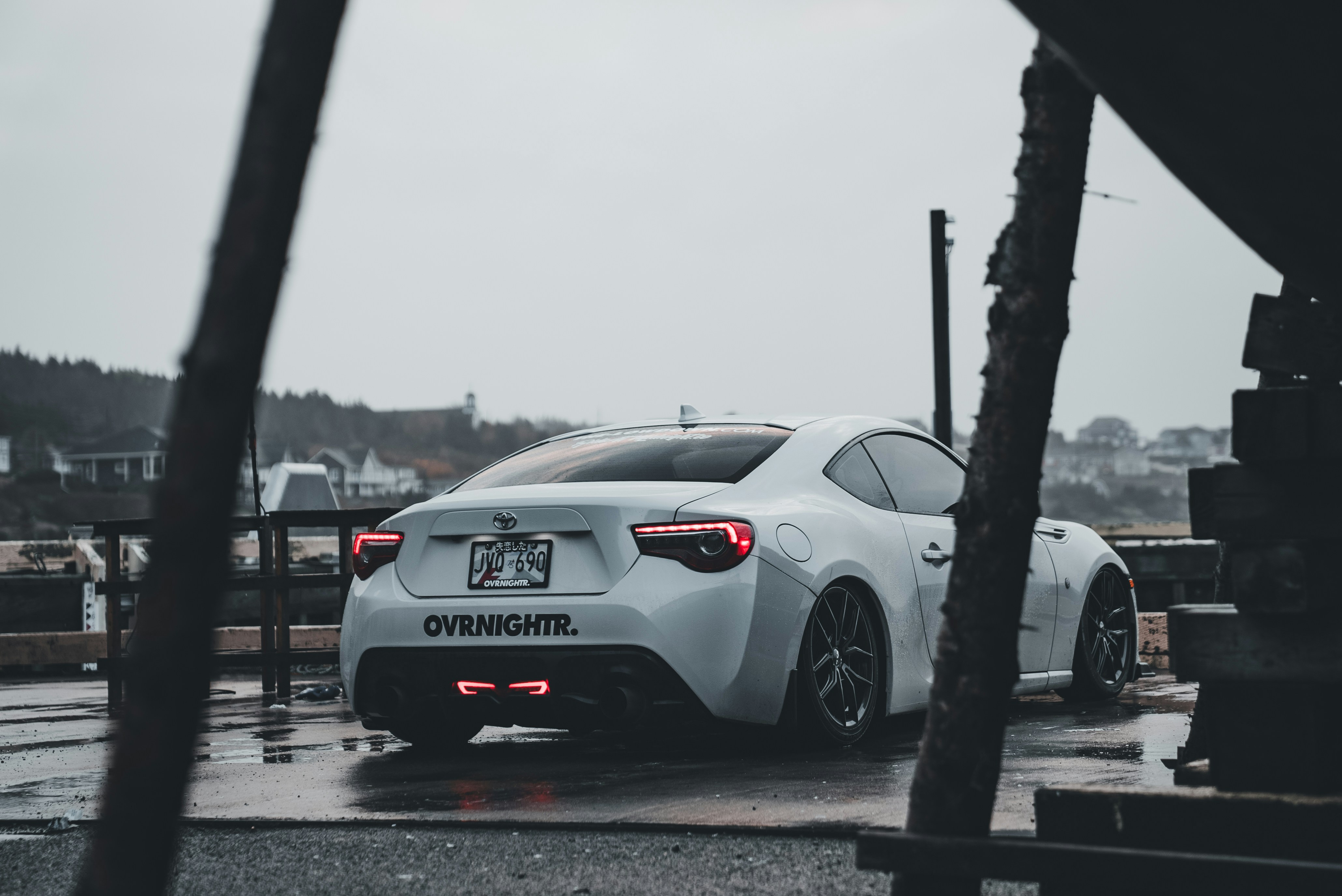 Rear-View Resilience: A White Sports Coupé Between Weathered Beams in the Rain