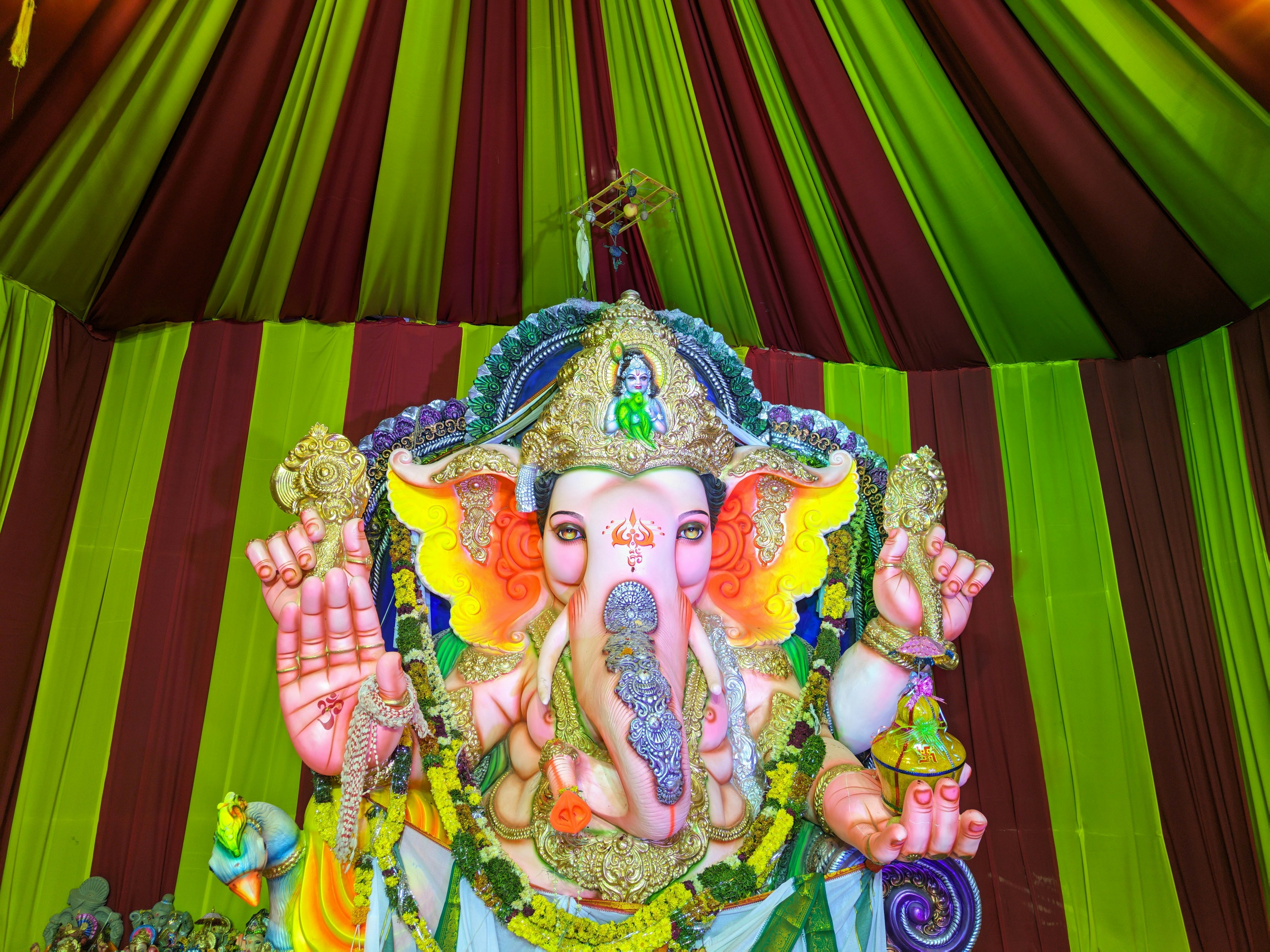 Photograph of a colorful Ganesha statue framed by a red and green striped canopy, captured in a festive shrine scene. The statue remains the focal point surrounded by vibrant fabric and decorative lighting.