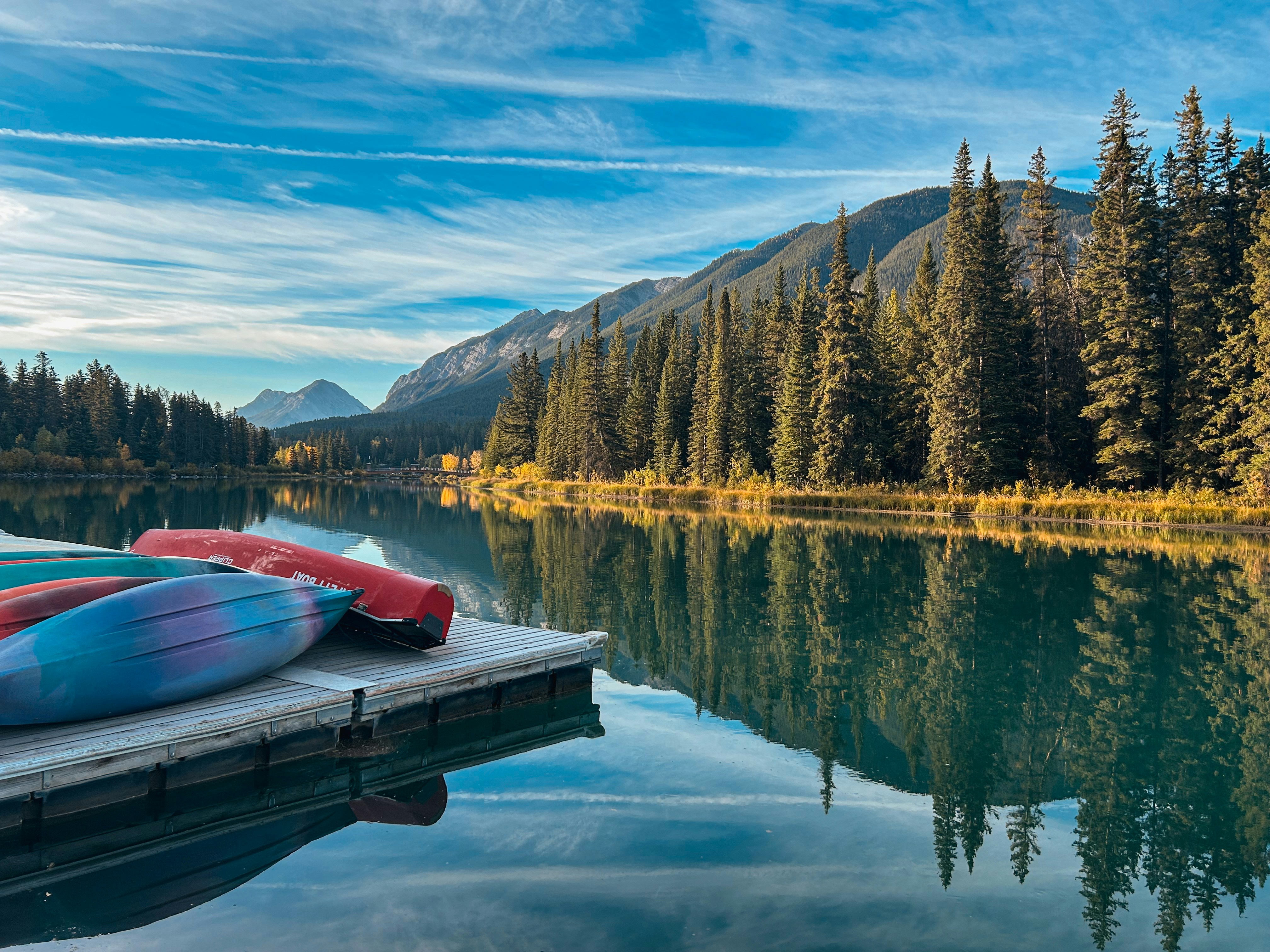 Canoes rest on a dock by a tranquil lake reflecting pine-covered mountains and a clear sky.