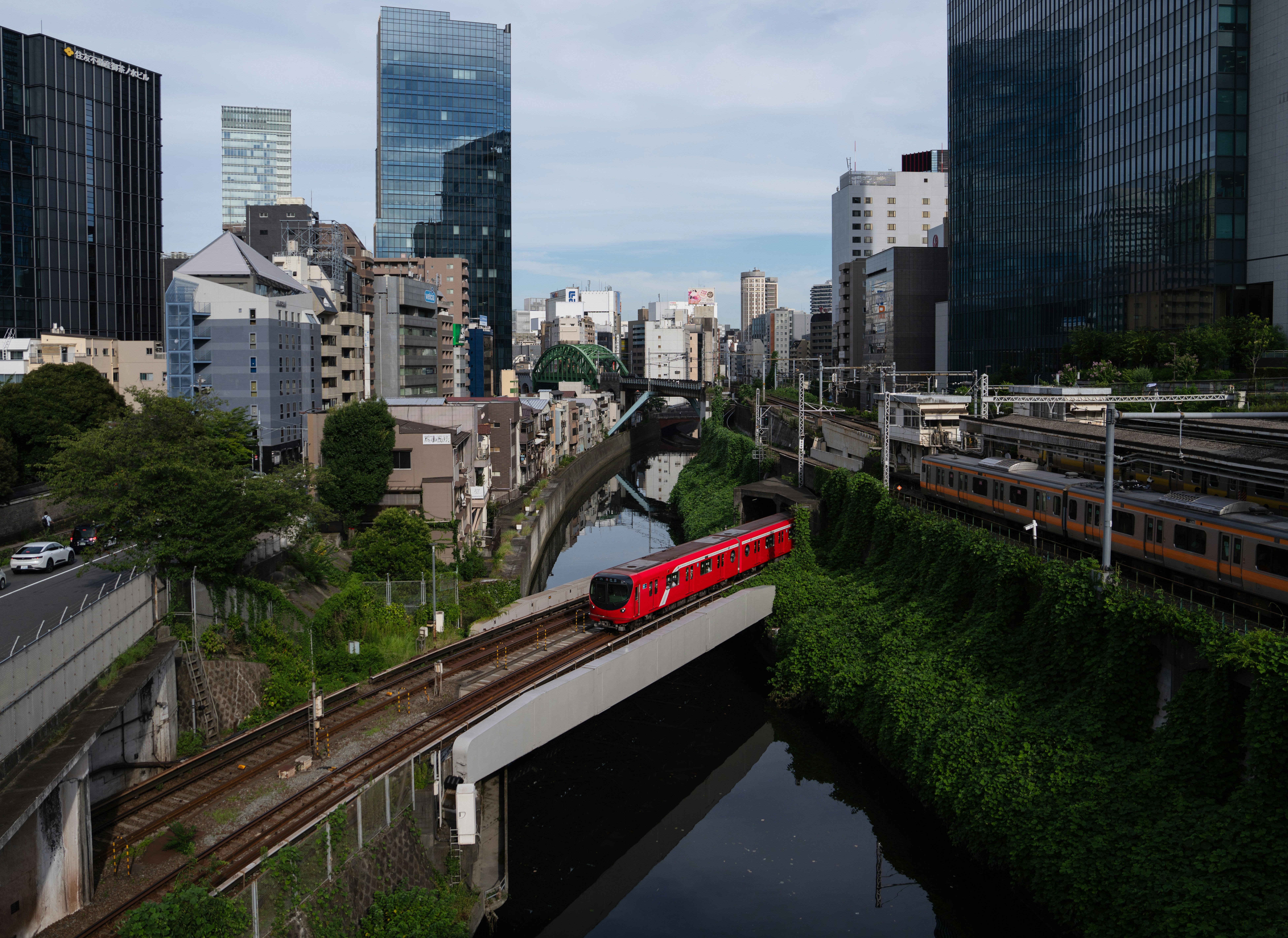 A vibrant red train glides over a lush, vine-covered bridge in the heart of Tokyo's bustling cityscape, reflecting the perfect blend of nature and modernity. (scene from Suzume no Tojimari)