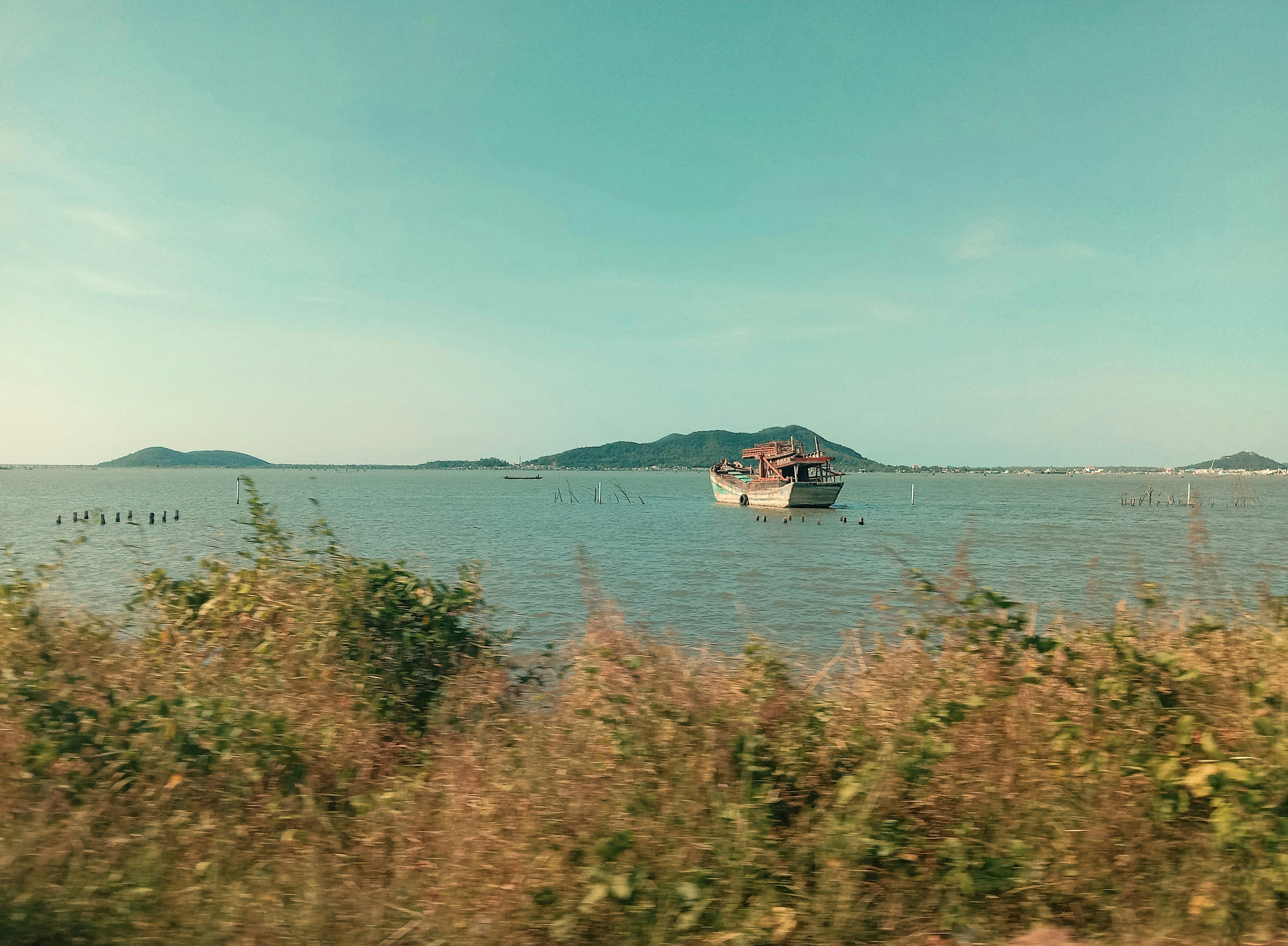 Calm sea with a lone boat near the horizon. Foreground grasses frame the scene against a clear blue sky.