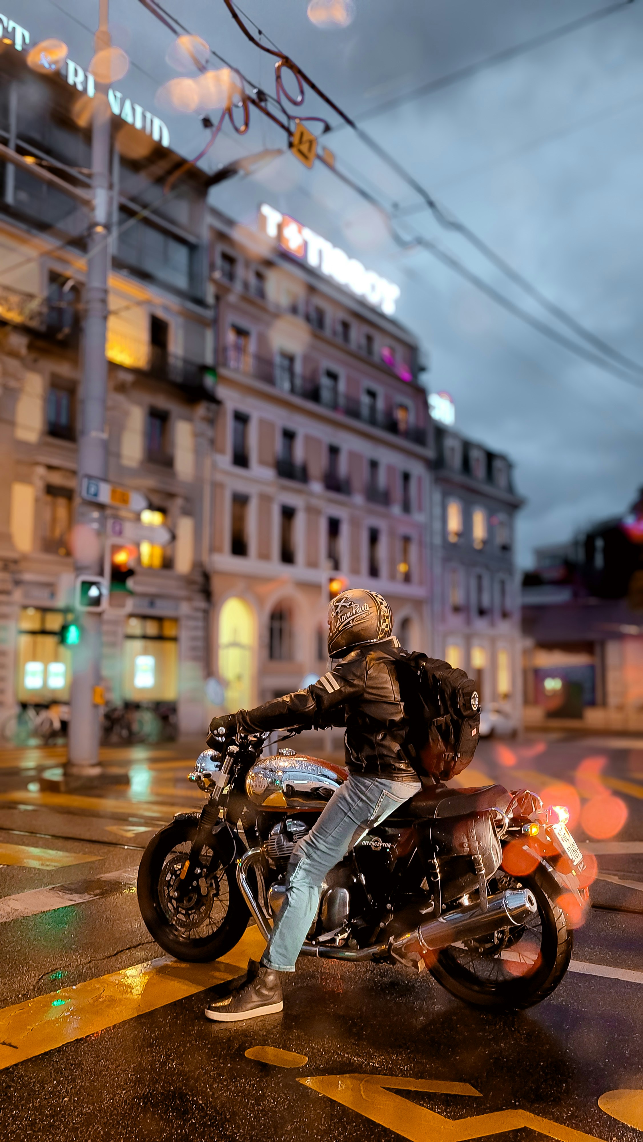 Motorcyclist in a helmet pauses at a city intersection, illuminated by streetlights and reflections on wet pavement.