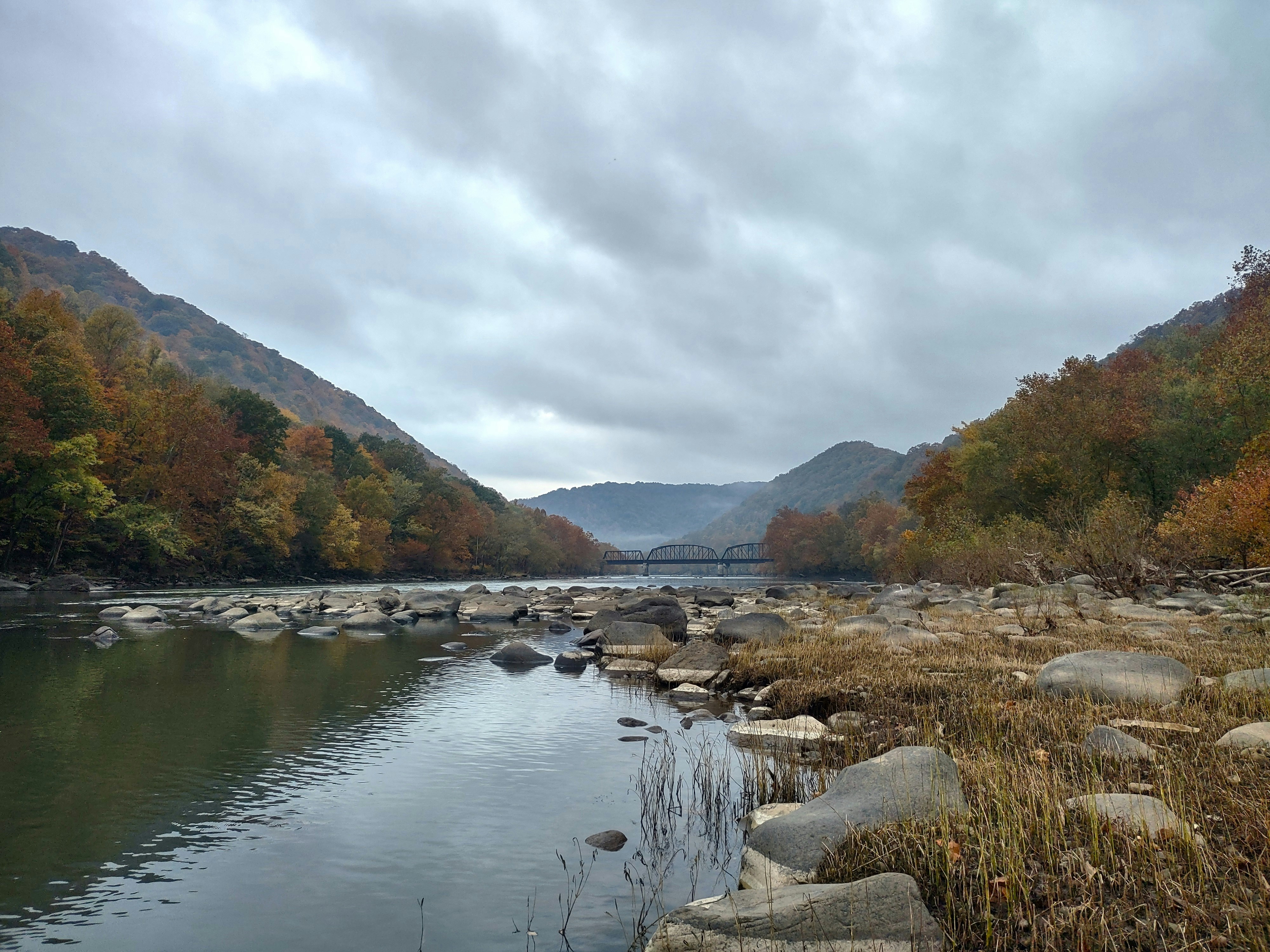 River winds through rocky banks lined with autumn foliage under a cloudy sky. Distant mountains stretch along the horizon, completing the tranquil landscape.