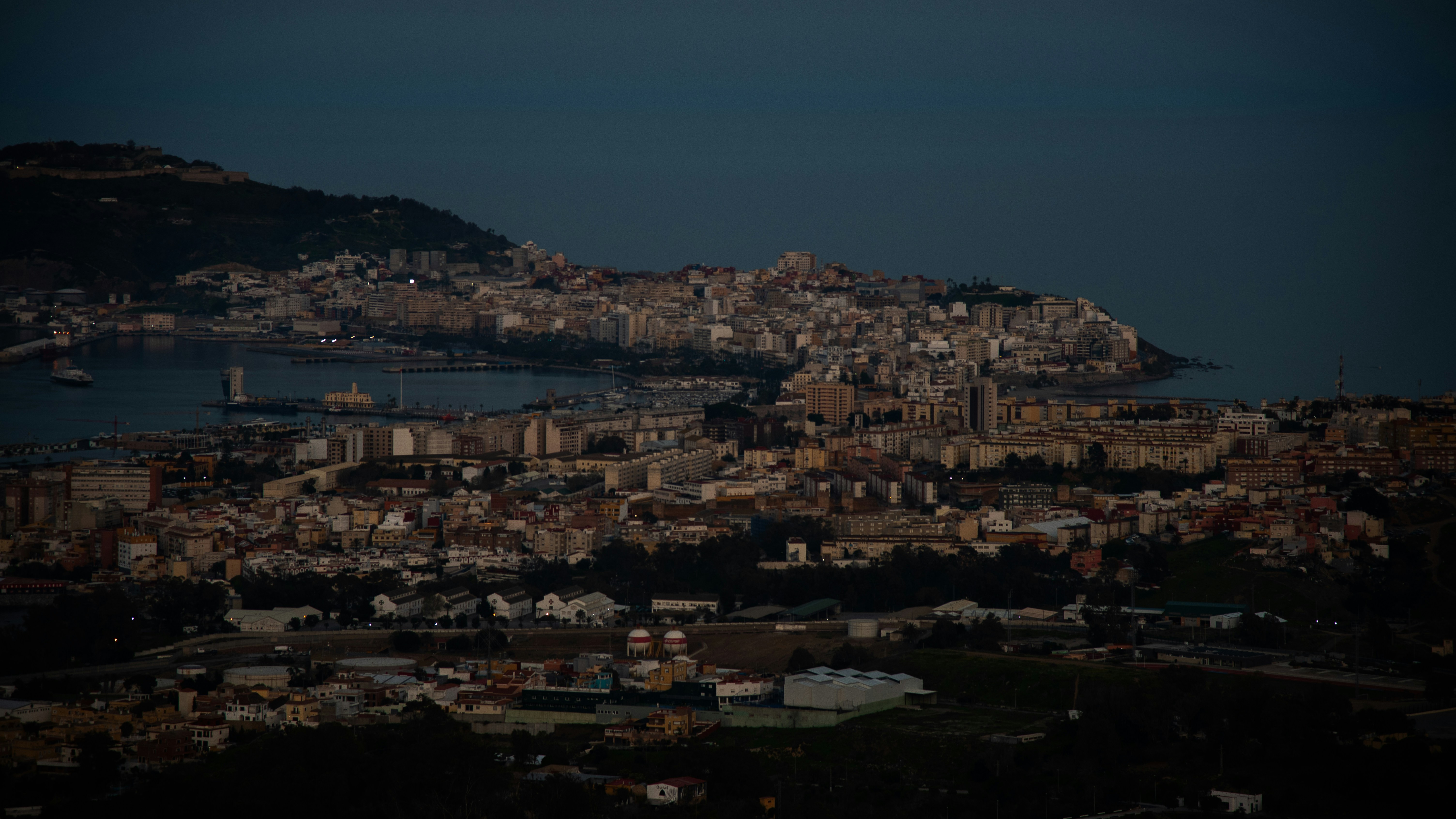 Distant view of a coastal city under a darkening sky, with lights beginning to twinkle along the shoreline.