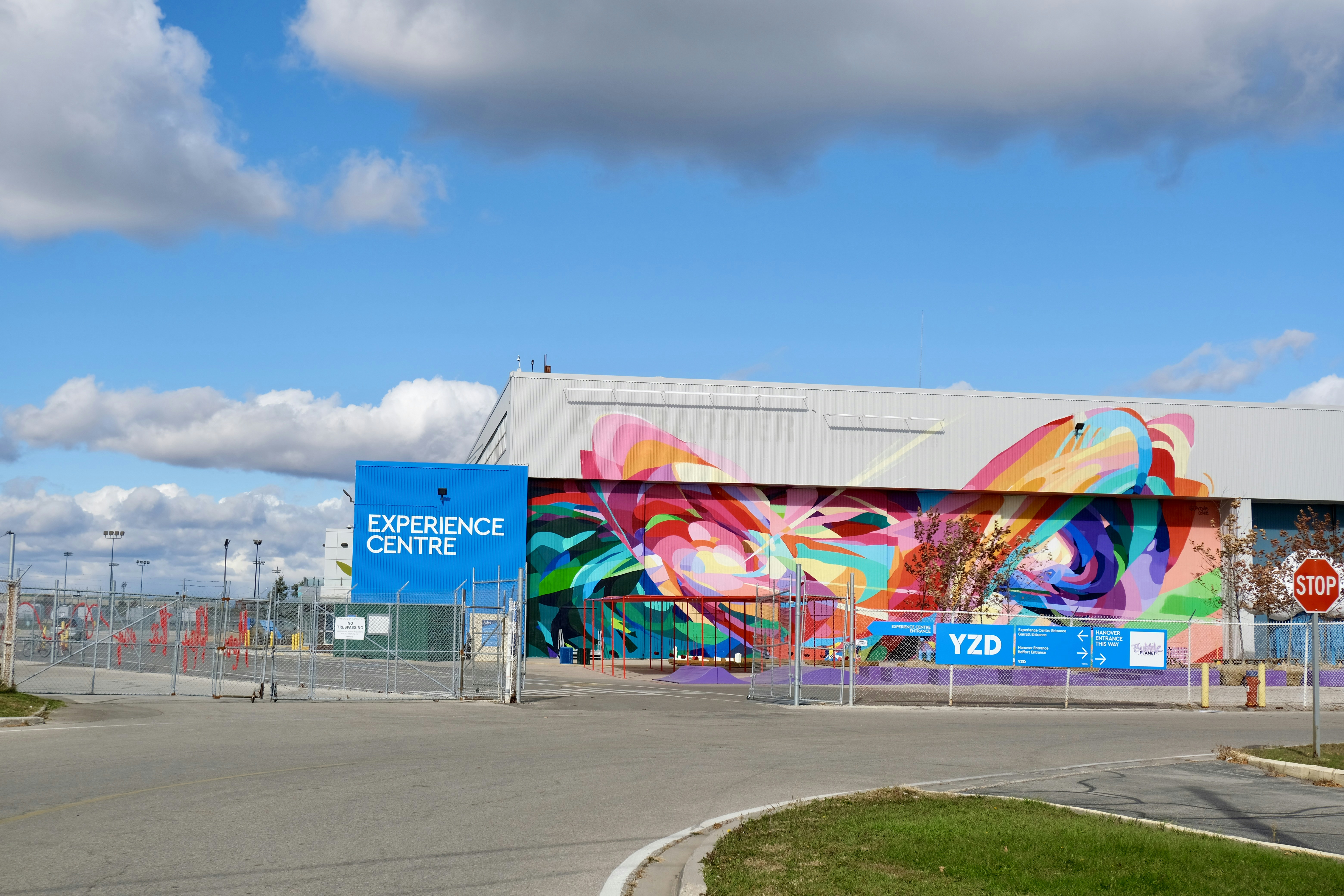 Colorful mural on an industrial building behind a fenced area under a bright blue sky.