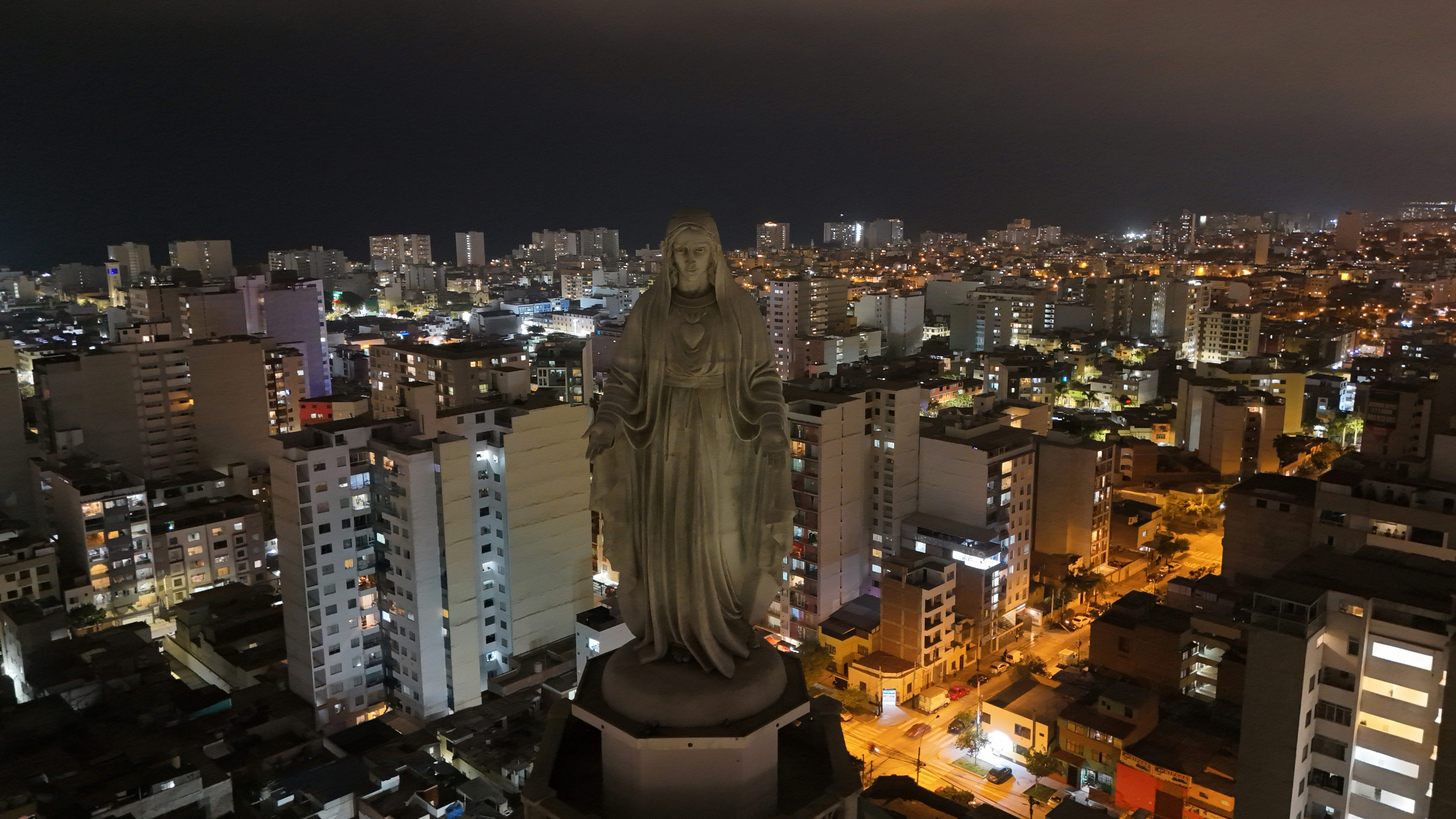 Night view of a towering statue overlooking the illuminated cityscape of Lima, Peru.