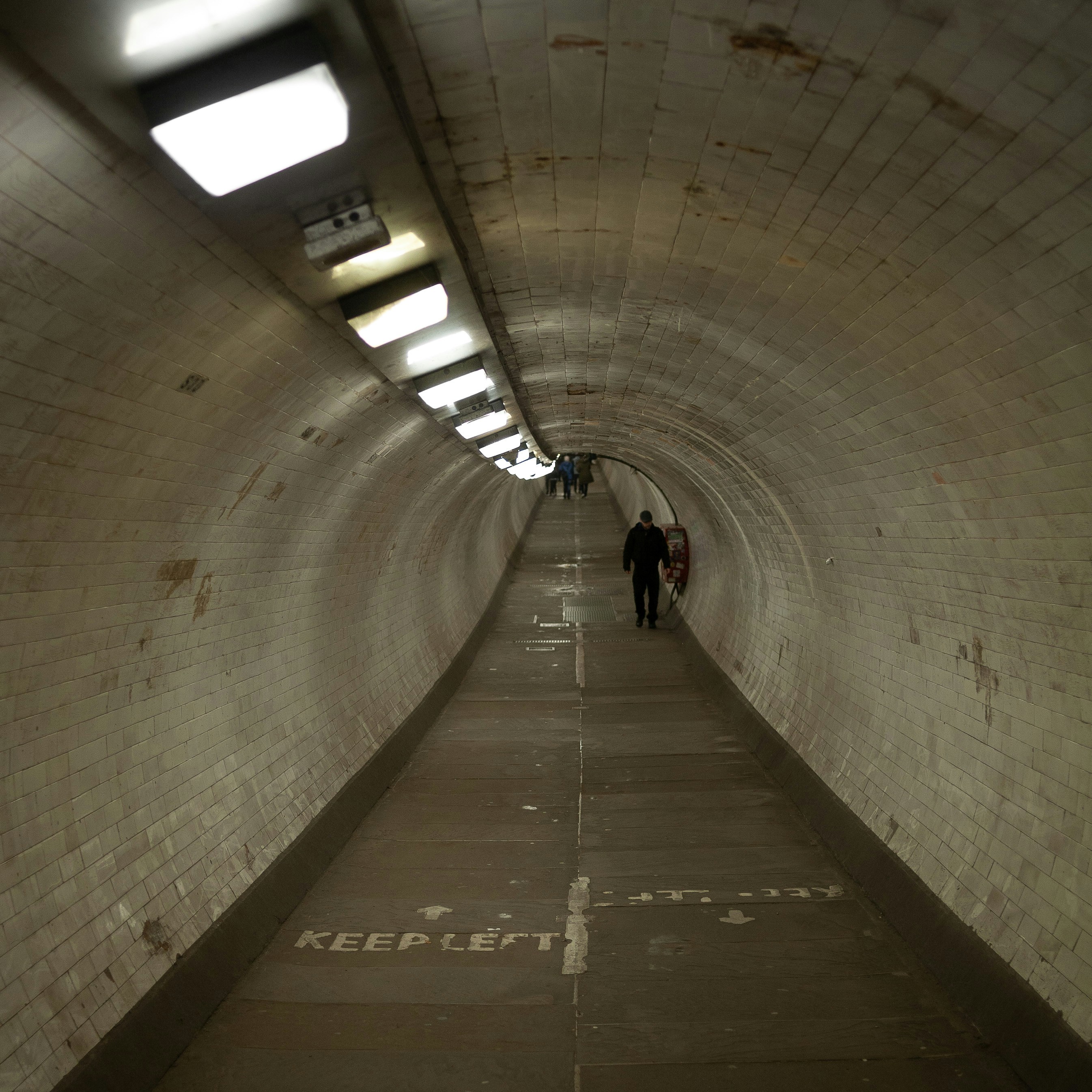 Pedestrian tunnel with brick walls, overhead lights, and a marked path with people walking.