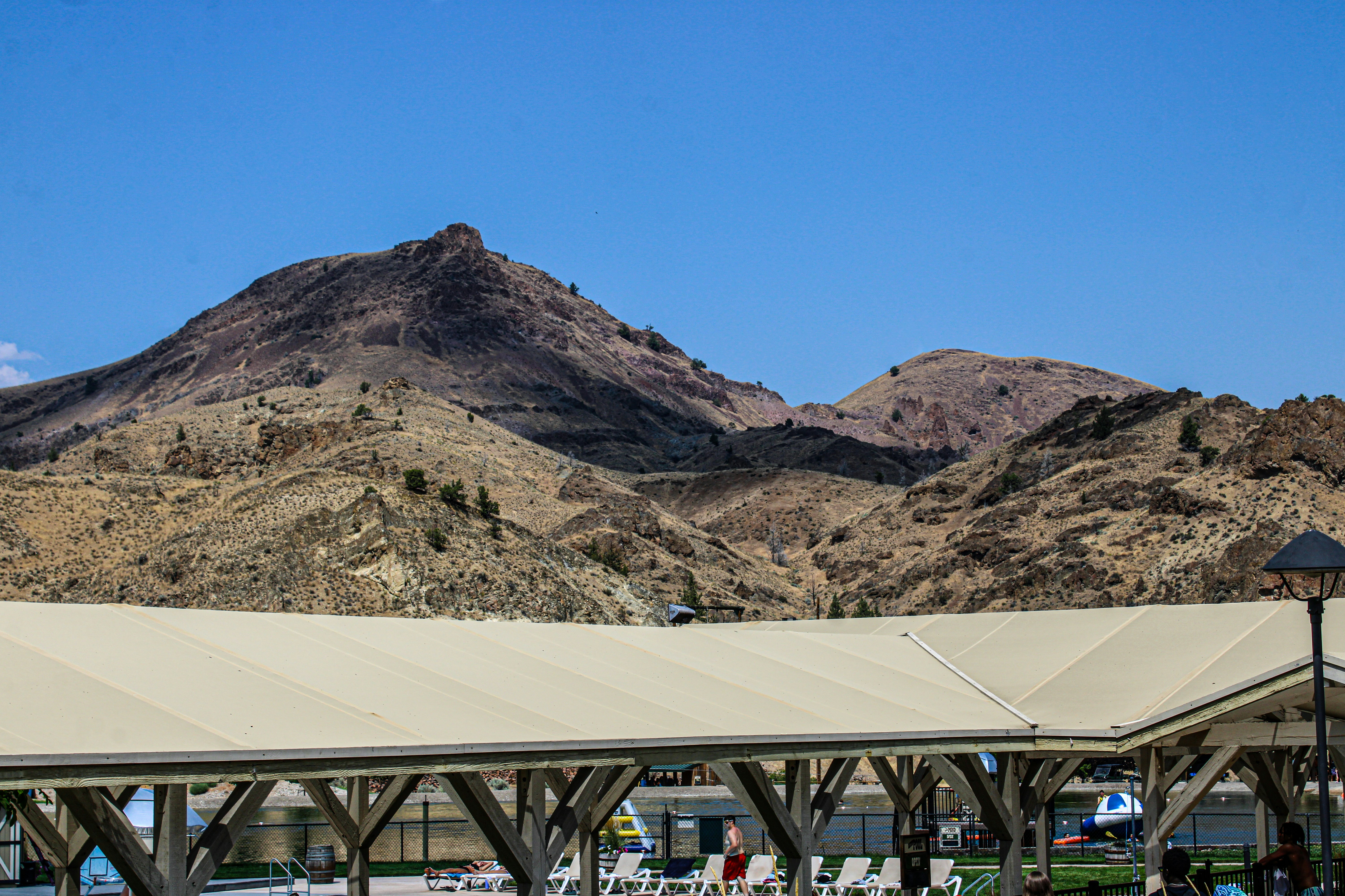 Desert mountains rise behind a long white tent-covered structure supported by wooden beams under a bright blue sky. The foreground shelter contrasts with the rugged terrain.