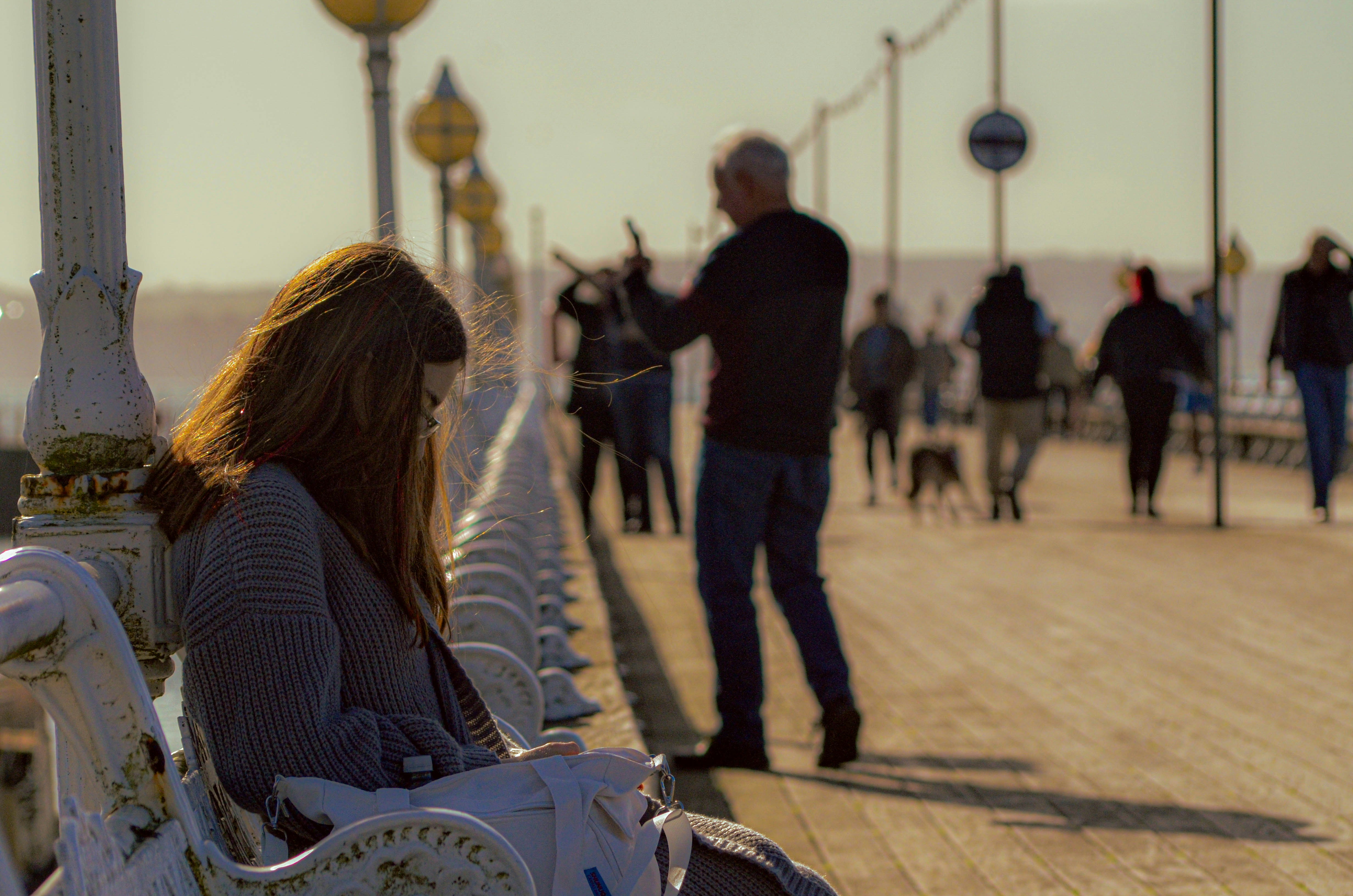 Woman sitting on a bench along a busy boardwalk, with people walking and a distant shoreline in the background.