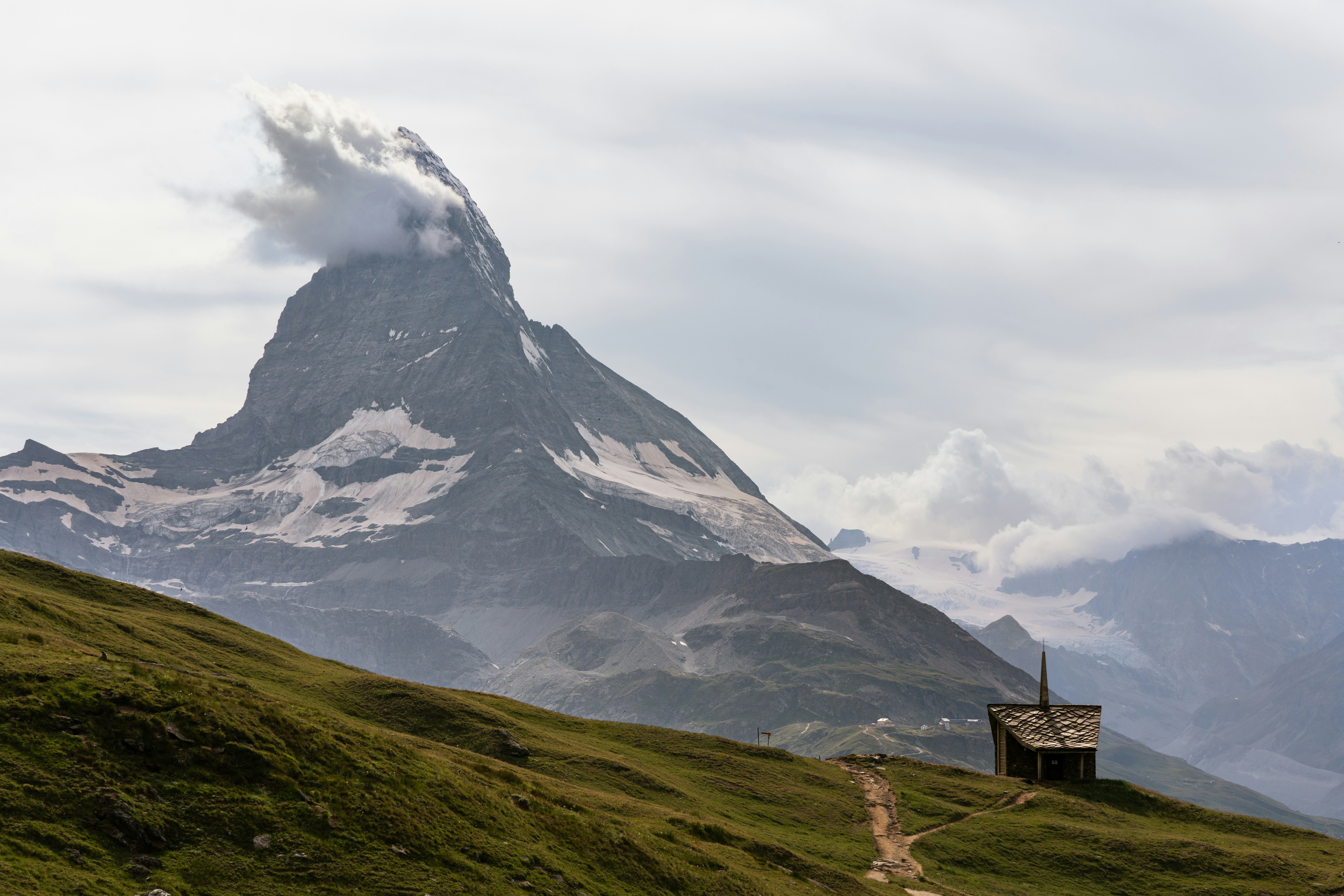 Mountain peak partially veiled by clouds with a small cabin in the foreground on a grassy hill.