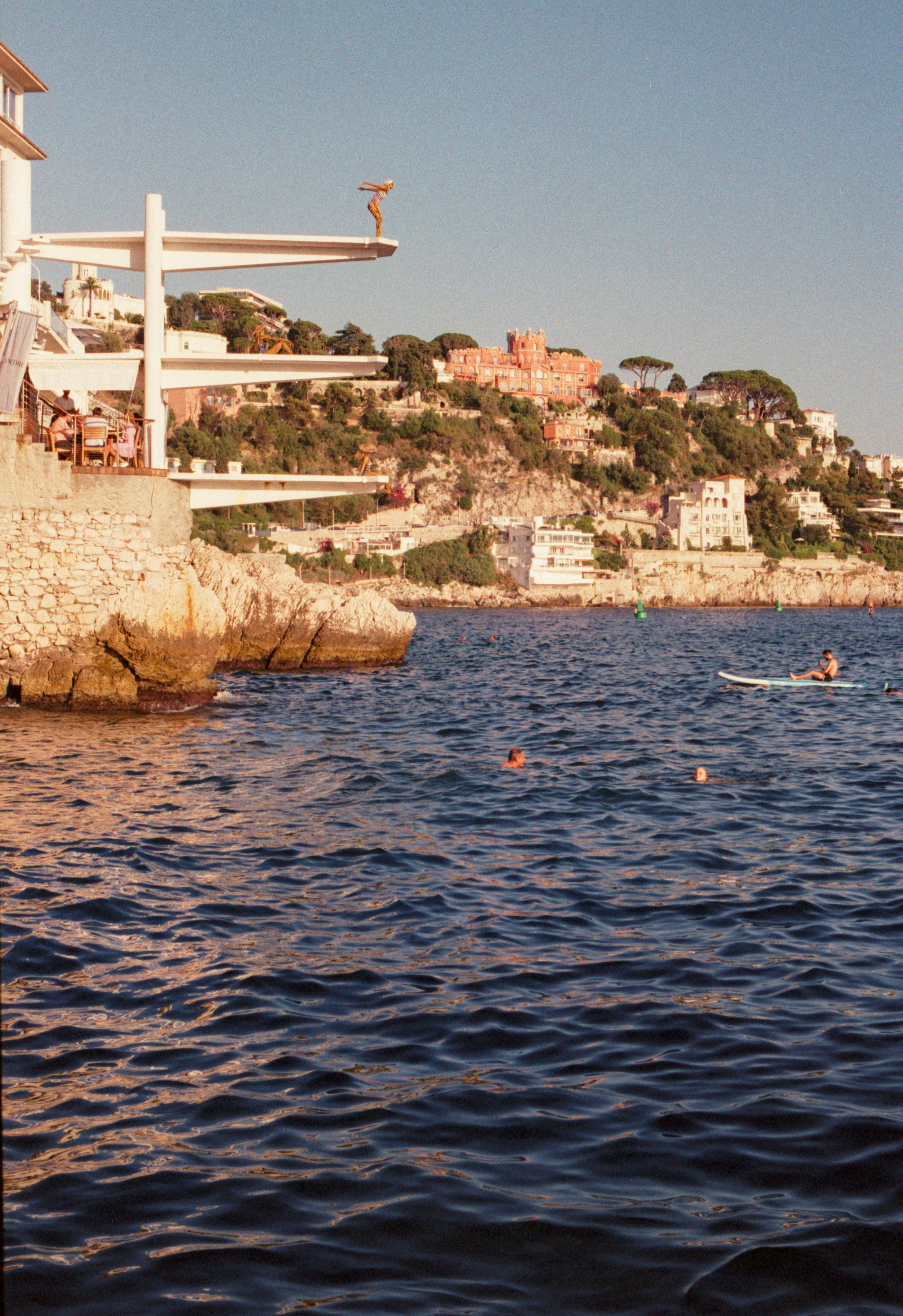 Swimmers enjoying a coastal view with rocky cliffs and distant hillside buildings at sunset.
