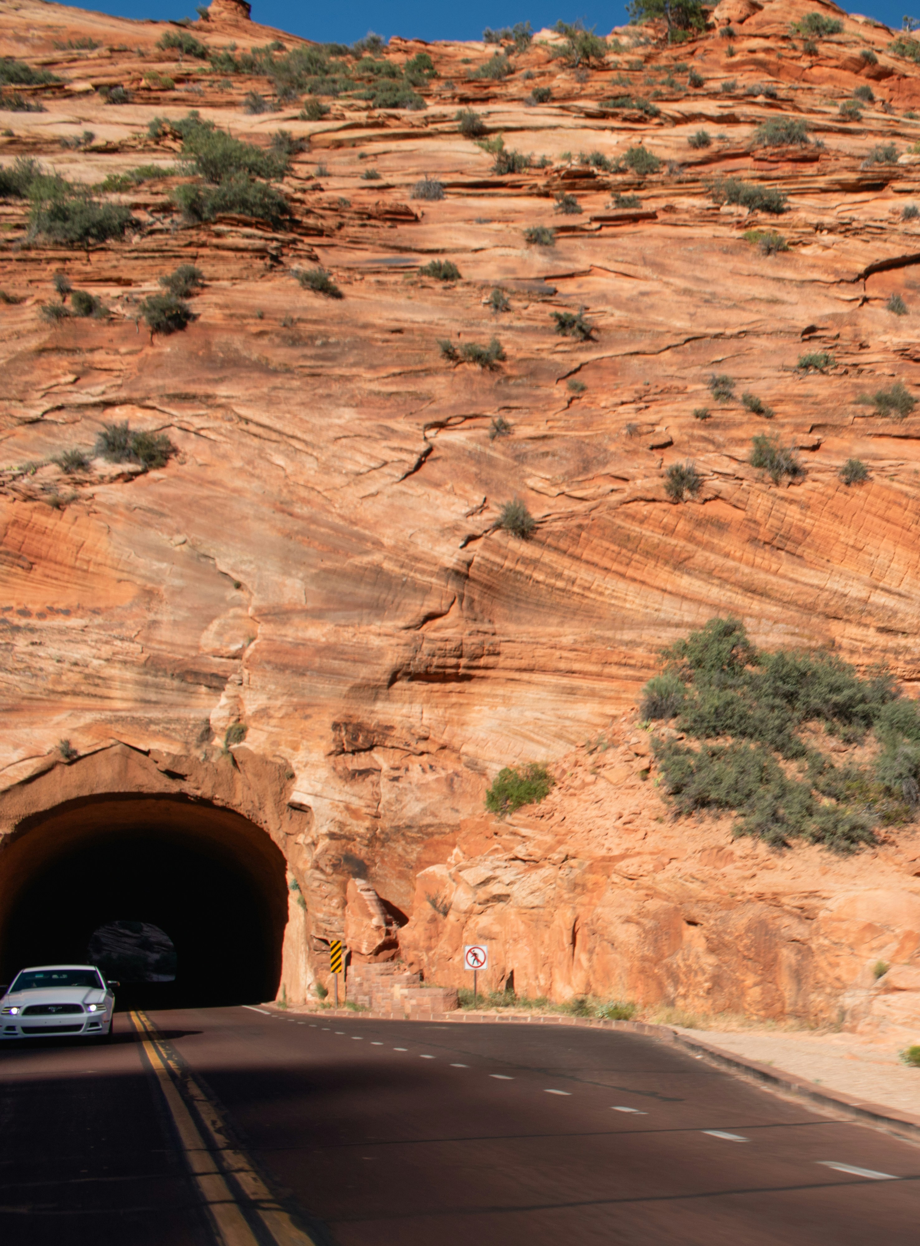 A sleek white car approaches a tunnel carved into the vibrant red rock of a canyon, framed by a clear blue sky above.