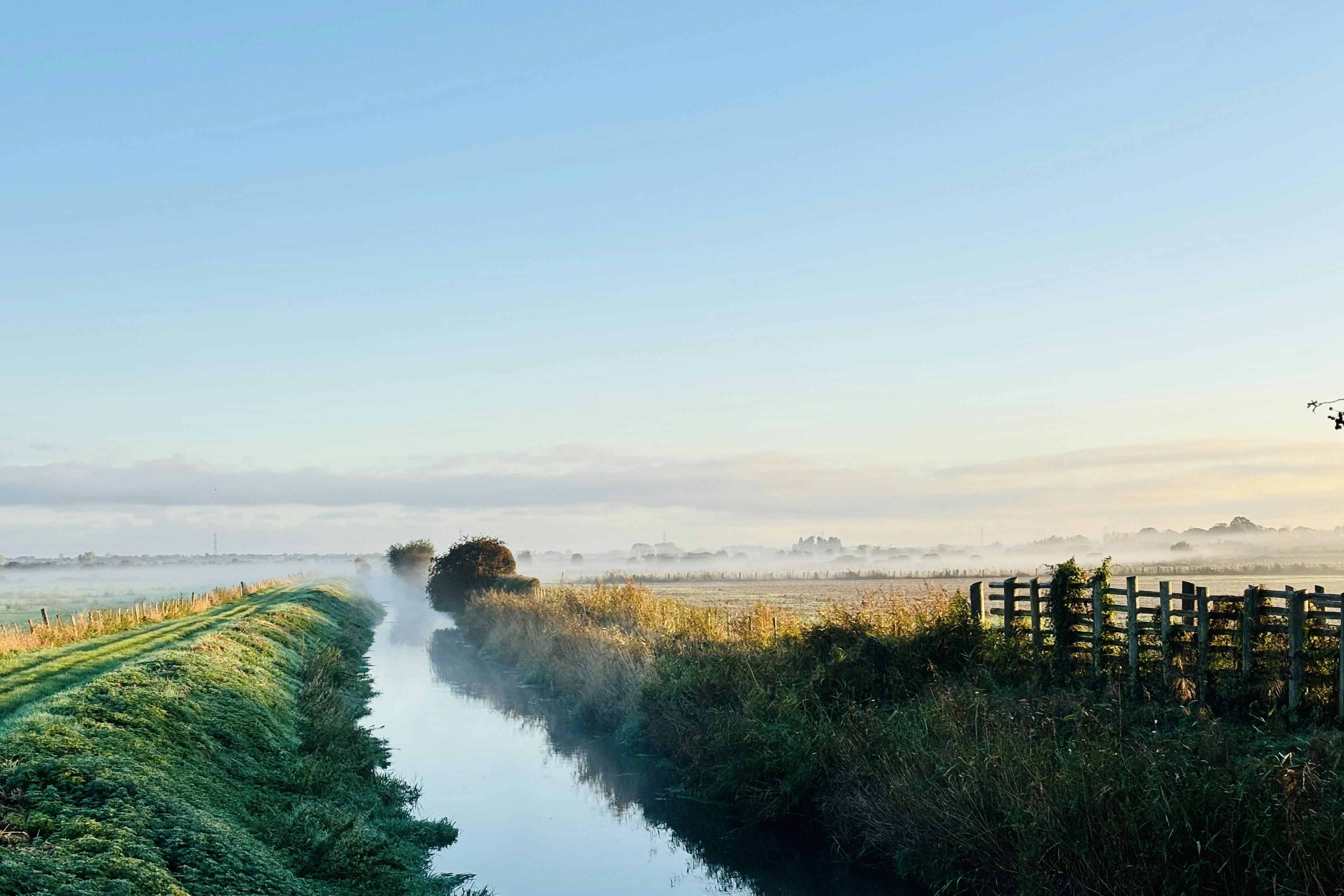 A tranquil dawn landscape featuring a narrow canal flanked by green fields and a wooden fence, with soft mist hovering above the water.
