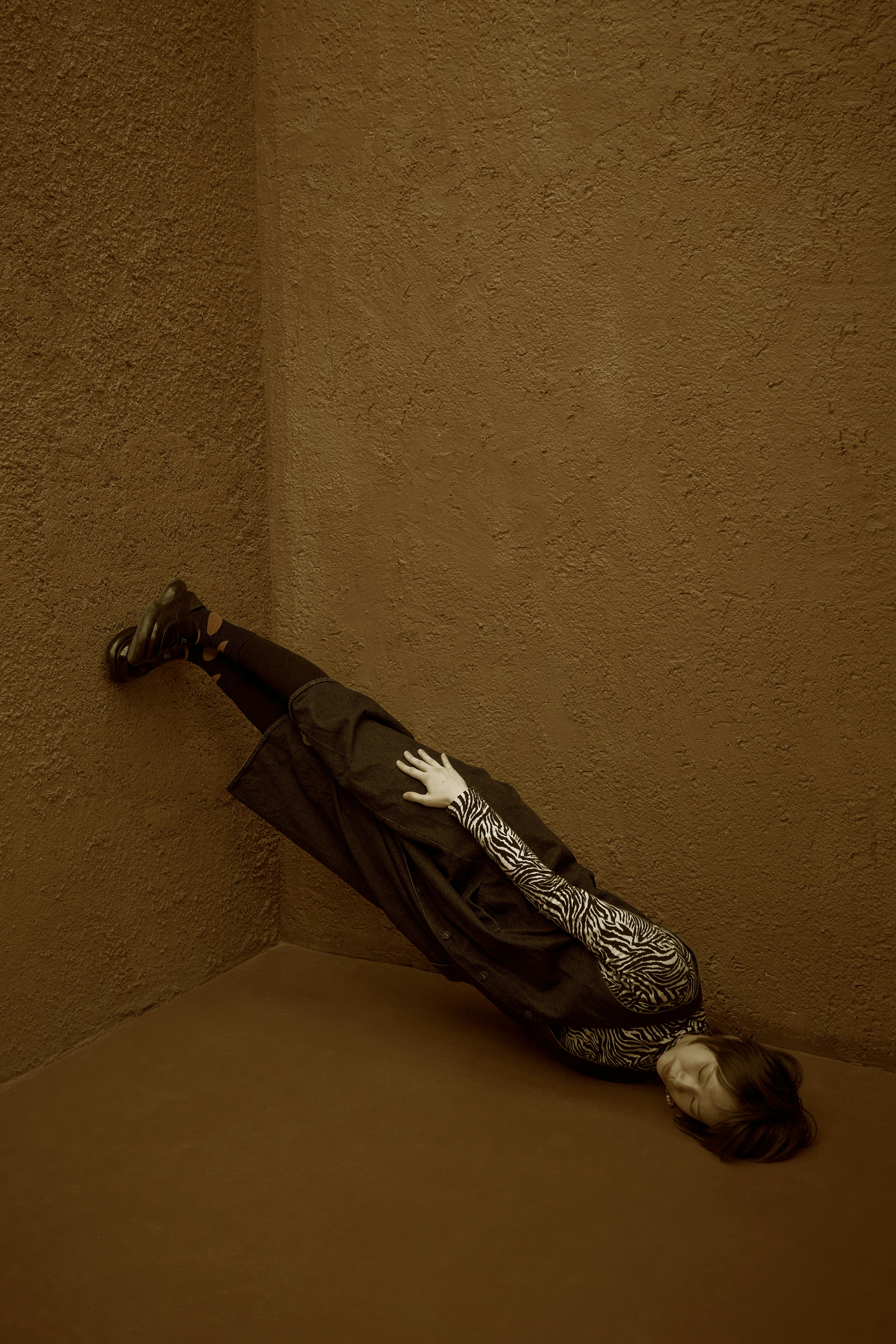 A figure in a black dress and striped sleeves leans against a textured brown wall, creating a surreal visual juxtaposition. The composition challenges perceptions of space and gravity.
