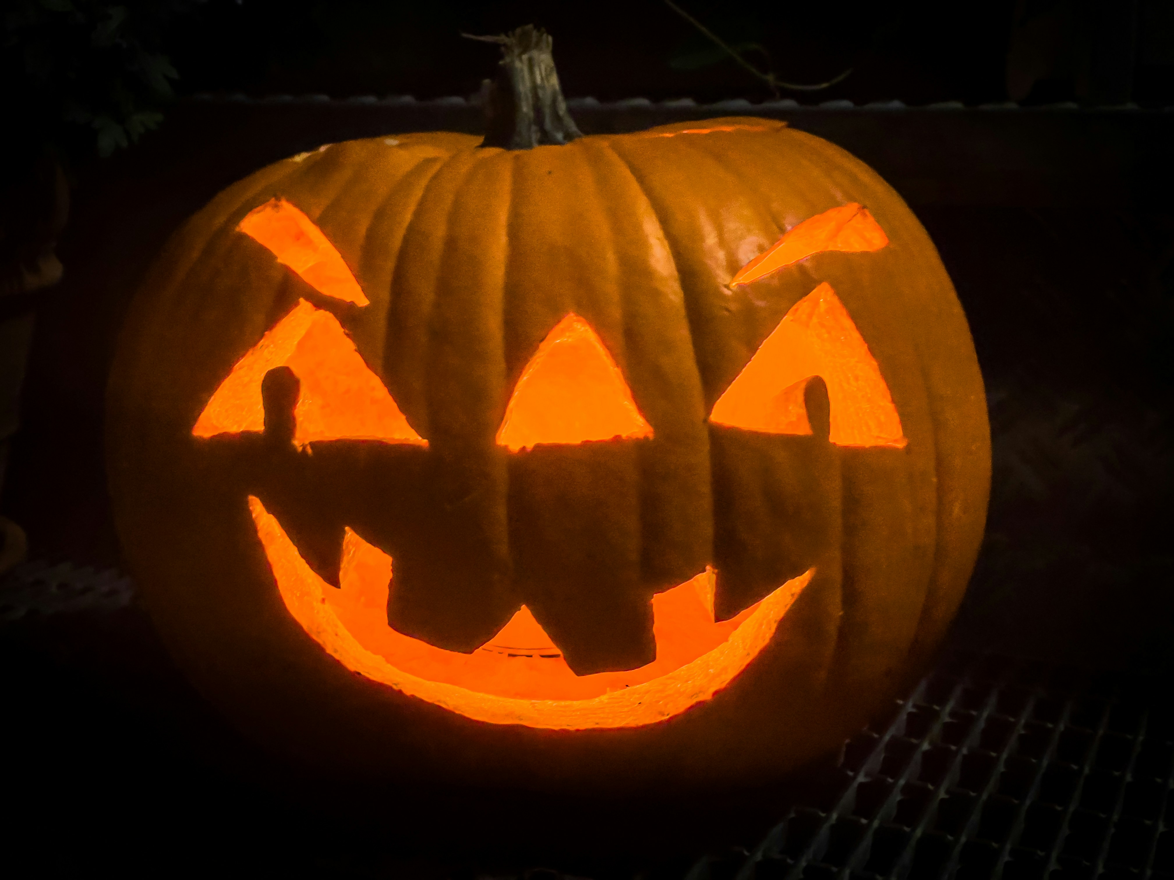 A single carved pumpkin with a cheerful, mischievous expression is lit from within, emitting a warm orange glow. The jack-o-lantern features triangle eyes, raised brows, and a wide, toothy grin, giving it a playful Halloween look. Set against a dark background, the pumpkin's light highlights its carved features, enhancing the festive Halloween atmosphere.