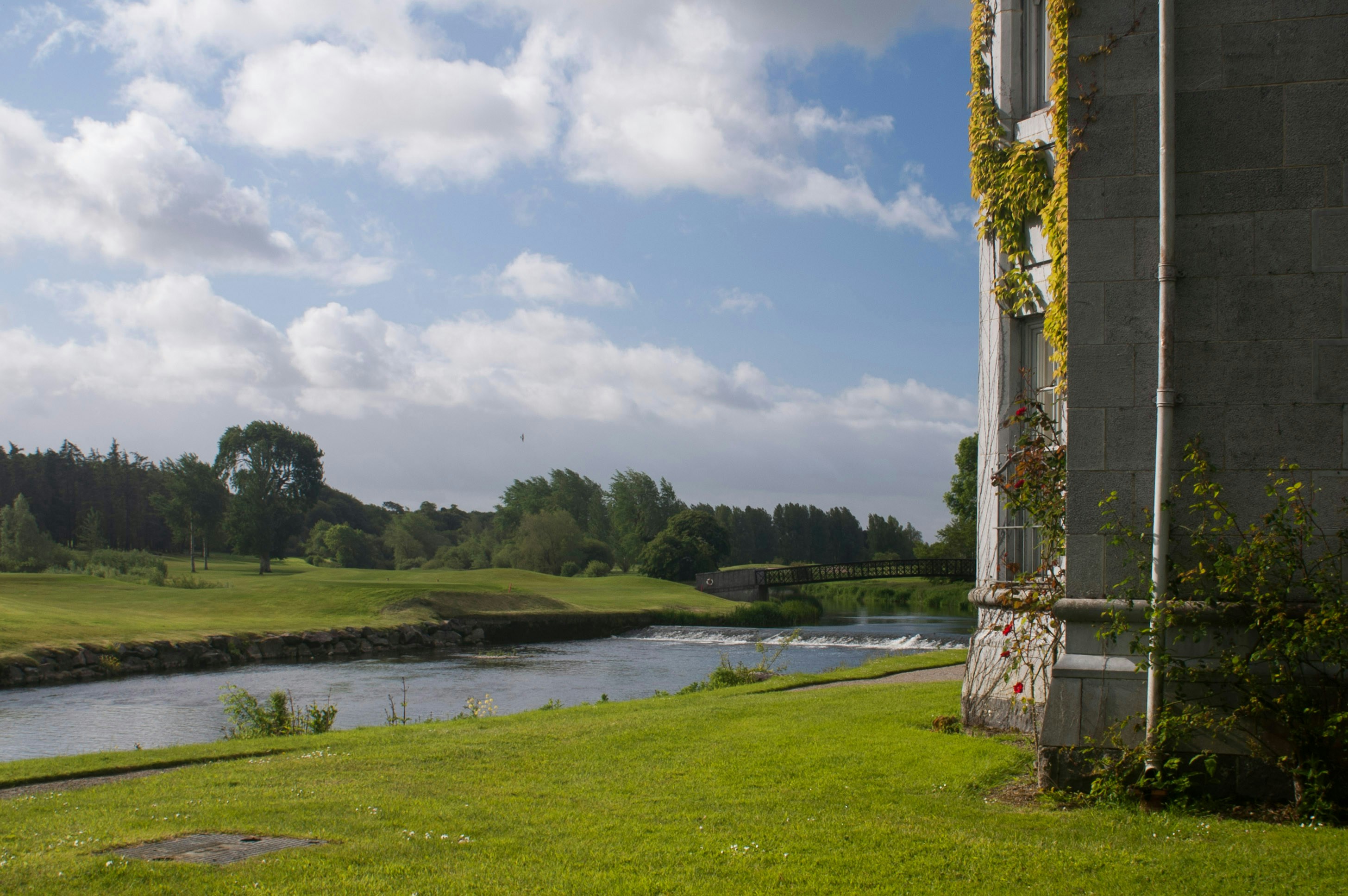 Sunlit riverside scene with an ivy-covered stone wall on the right, a calm river bend, and a grassy foreground under a partly cloudy sky.