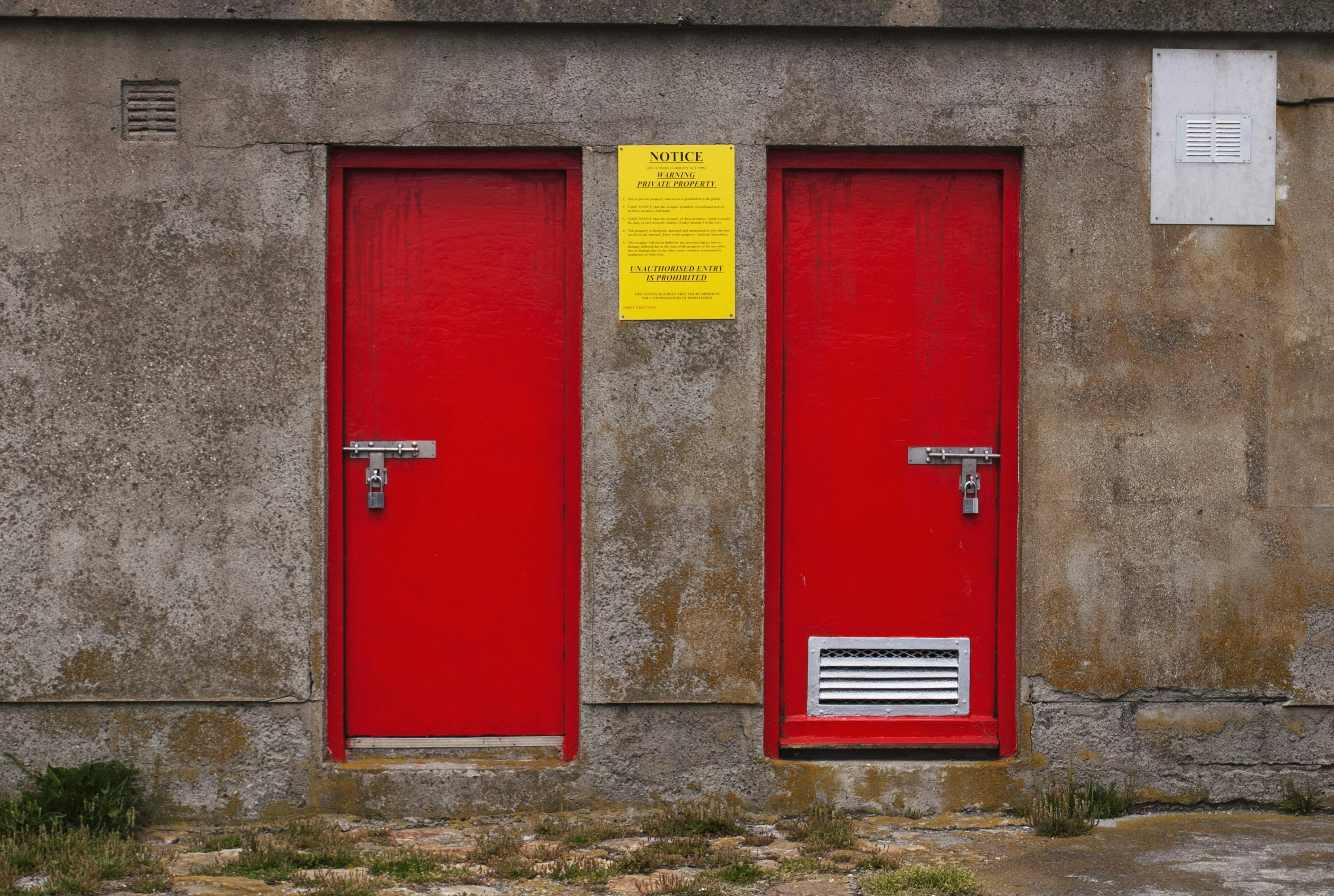Two bright red doors stand in a weathered concrete wall. A bold yellow warning notice sits between them, with a vent on the lower portion of the right door and rough, grassy ground in front.