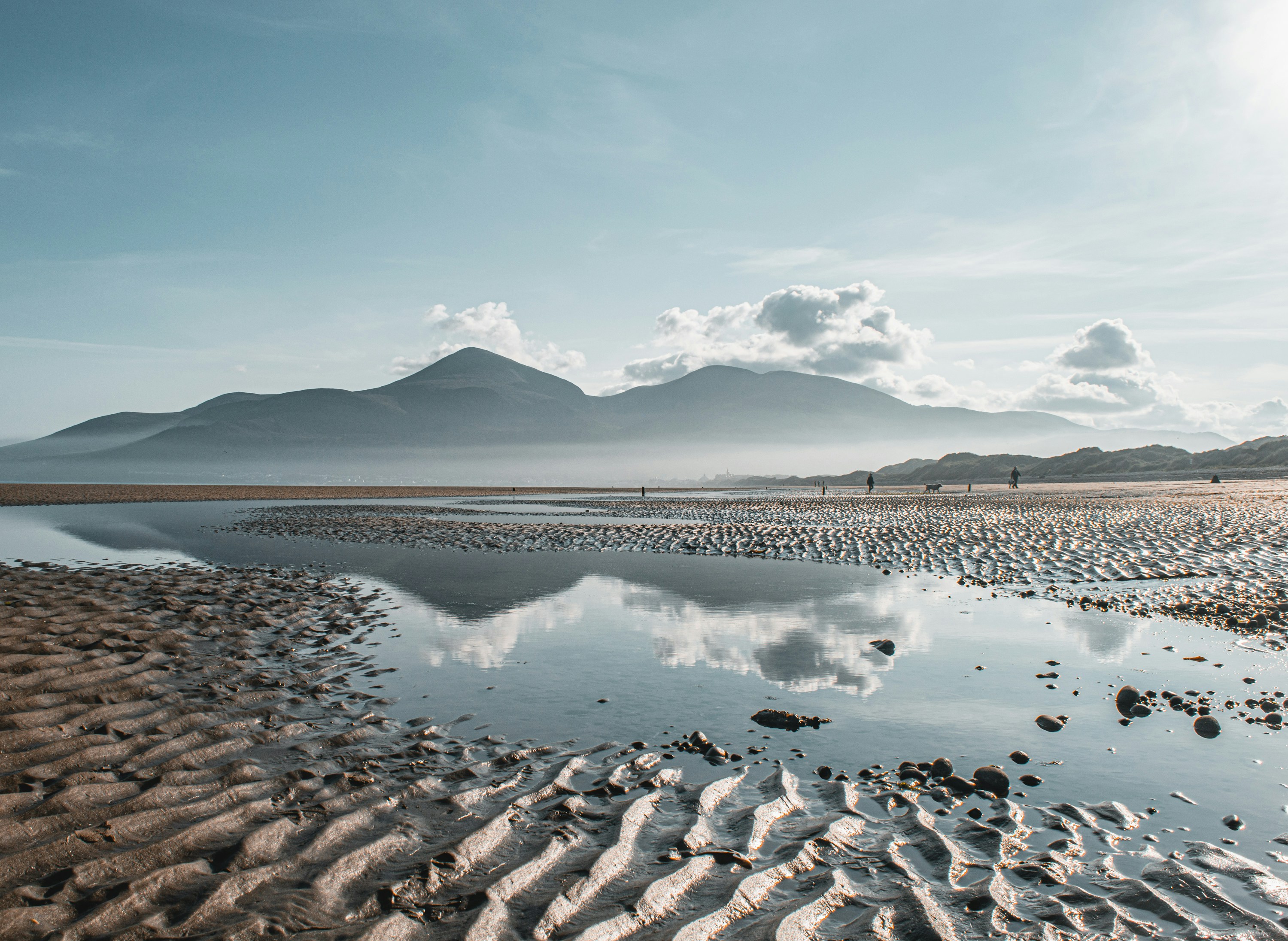 Rippling sand and a calm tidal pool mirror distant misty mountains under a soft sky.