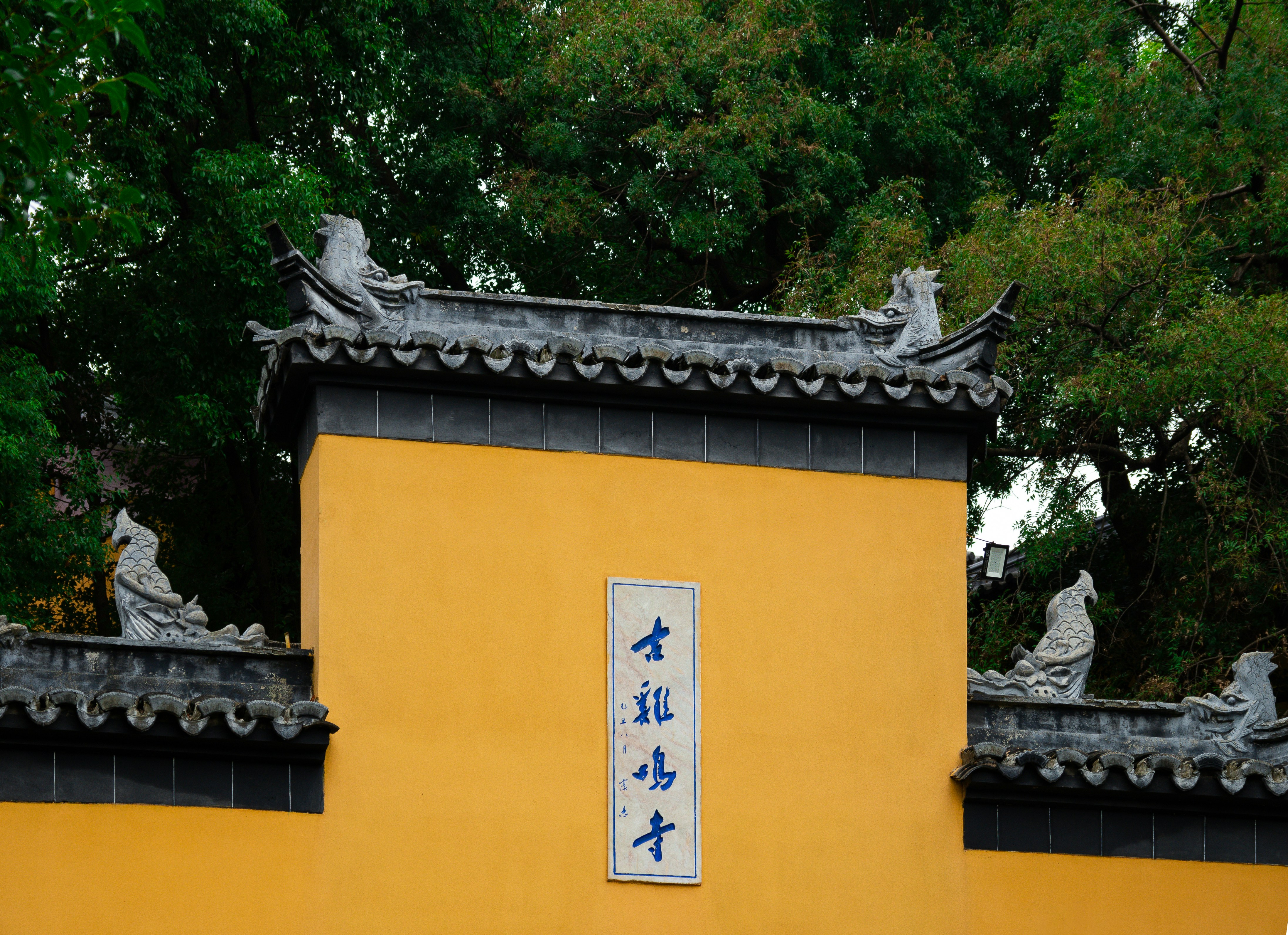 Yellow wall with a vertical blue inscription plaque, flanked by gray tiled eaves and lush green trees in the background.
