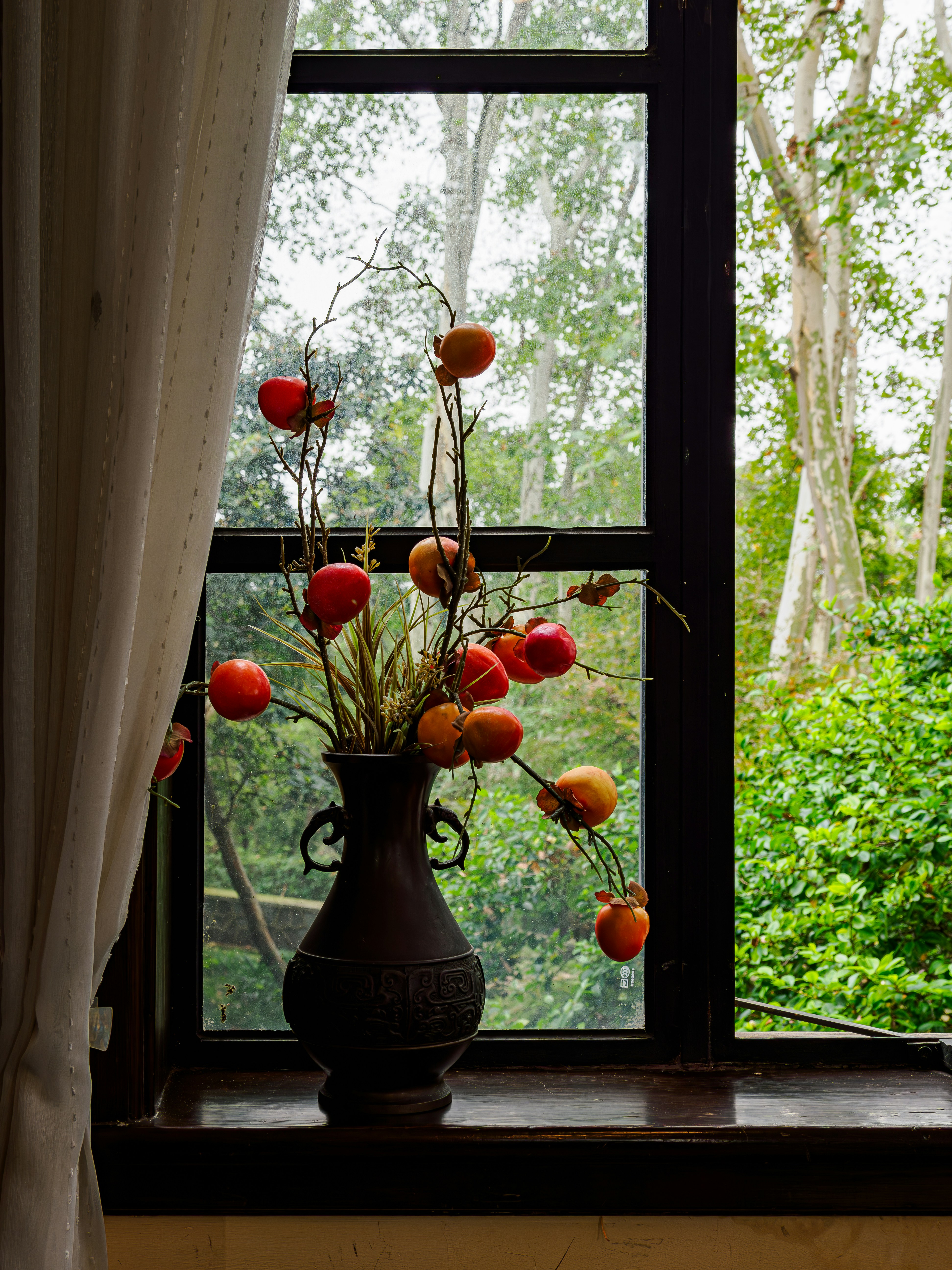Dark ceramic vase with bare-branch stems and red apples sits on a wooden windowsill. Sunlight from the window softens the scene, with green garden visible beyond.