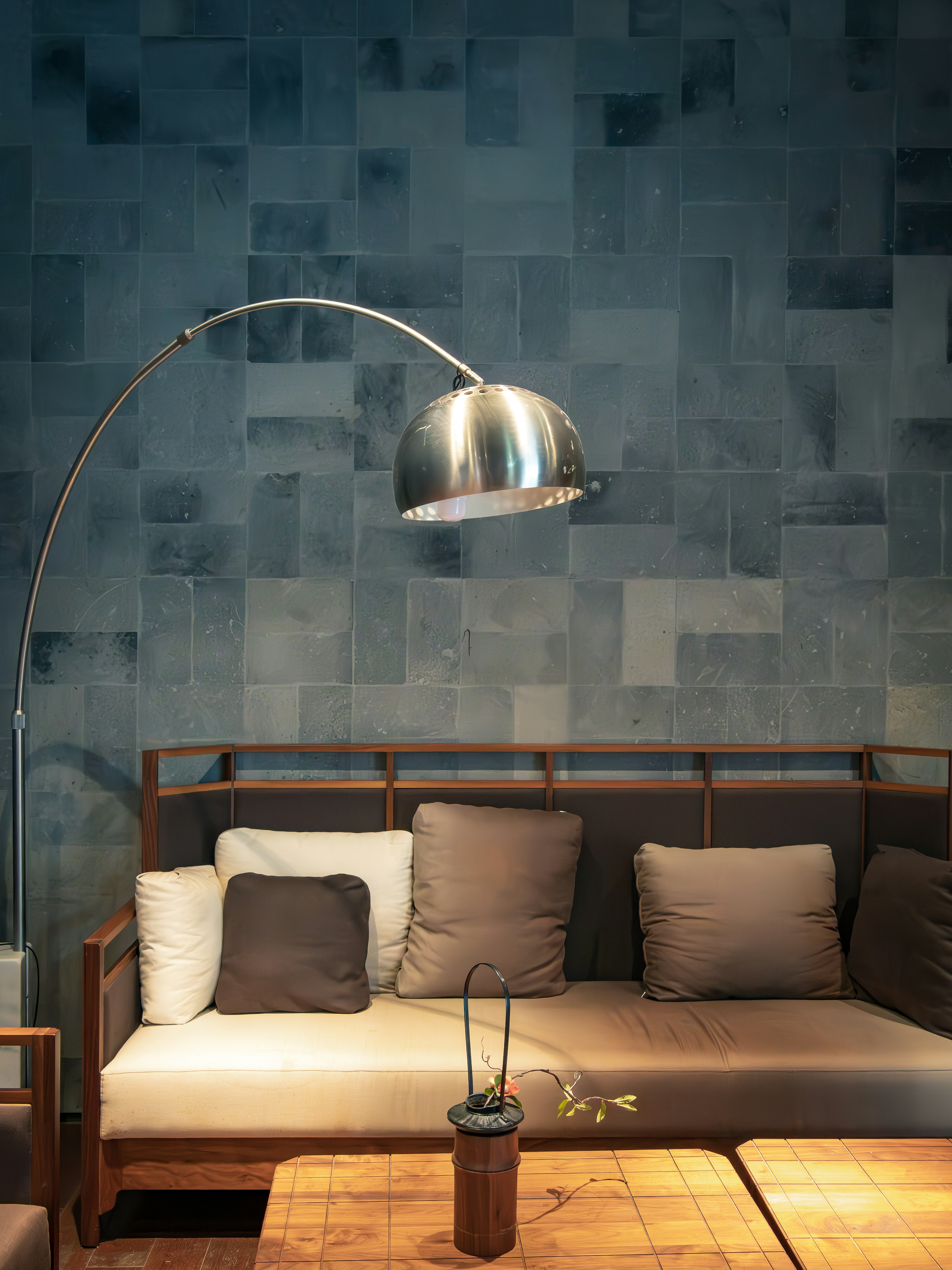 Chrome arc lamp arches over a cream sofa against a blue-tiled wall, with a wooden coffee table and a small plant in the foreground.