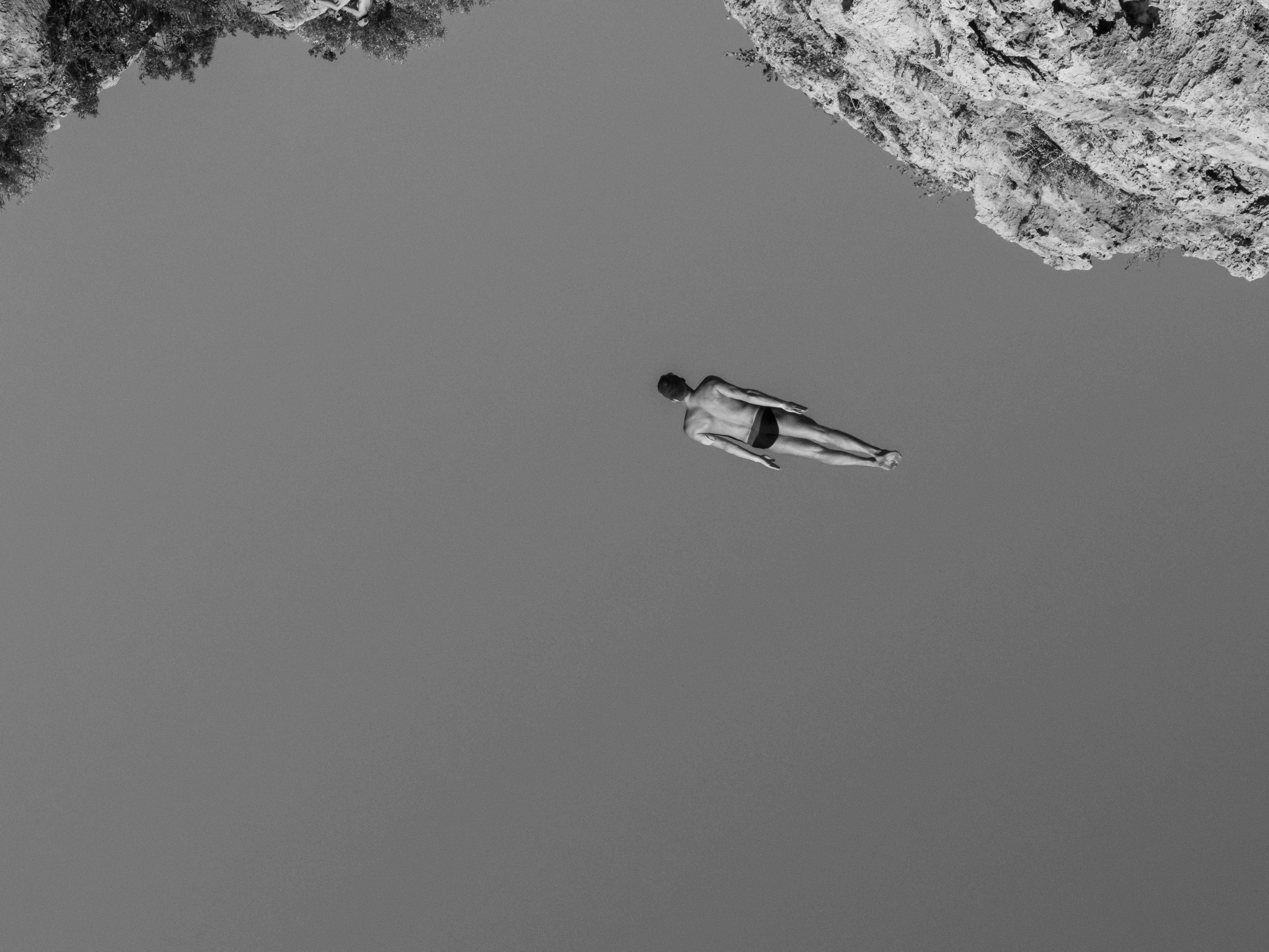 Minimalist photograph of a lone figure floating on flat gray water, framed by rugged cliff faces above. The scene emphasizes isolation and calm, rendered in a stark monochrome palette.