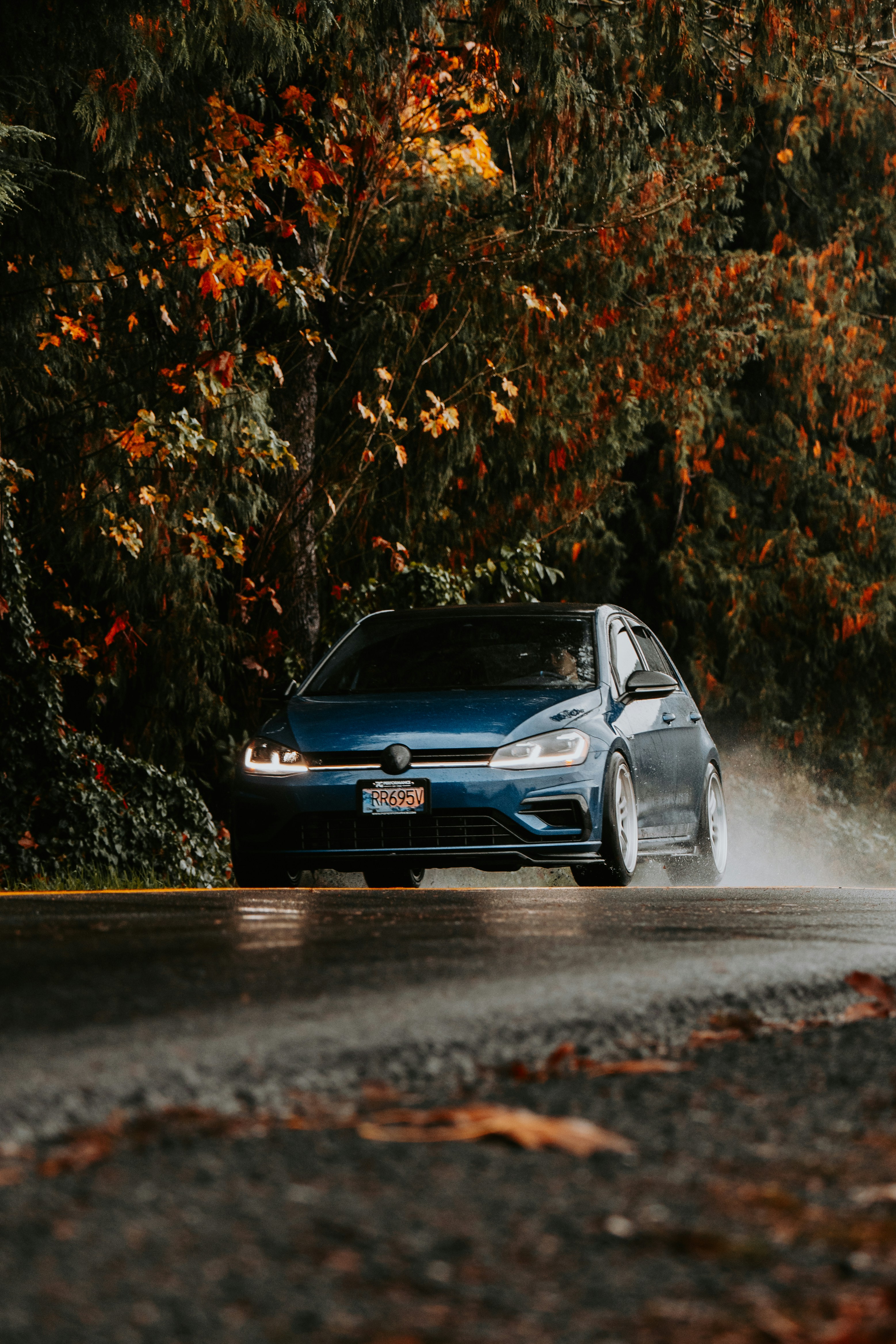 Blue sedan carves through autumn rain on a leaf-strewn, wet forest road