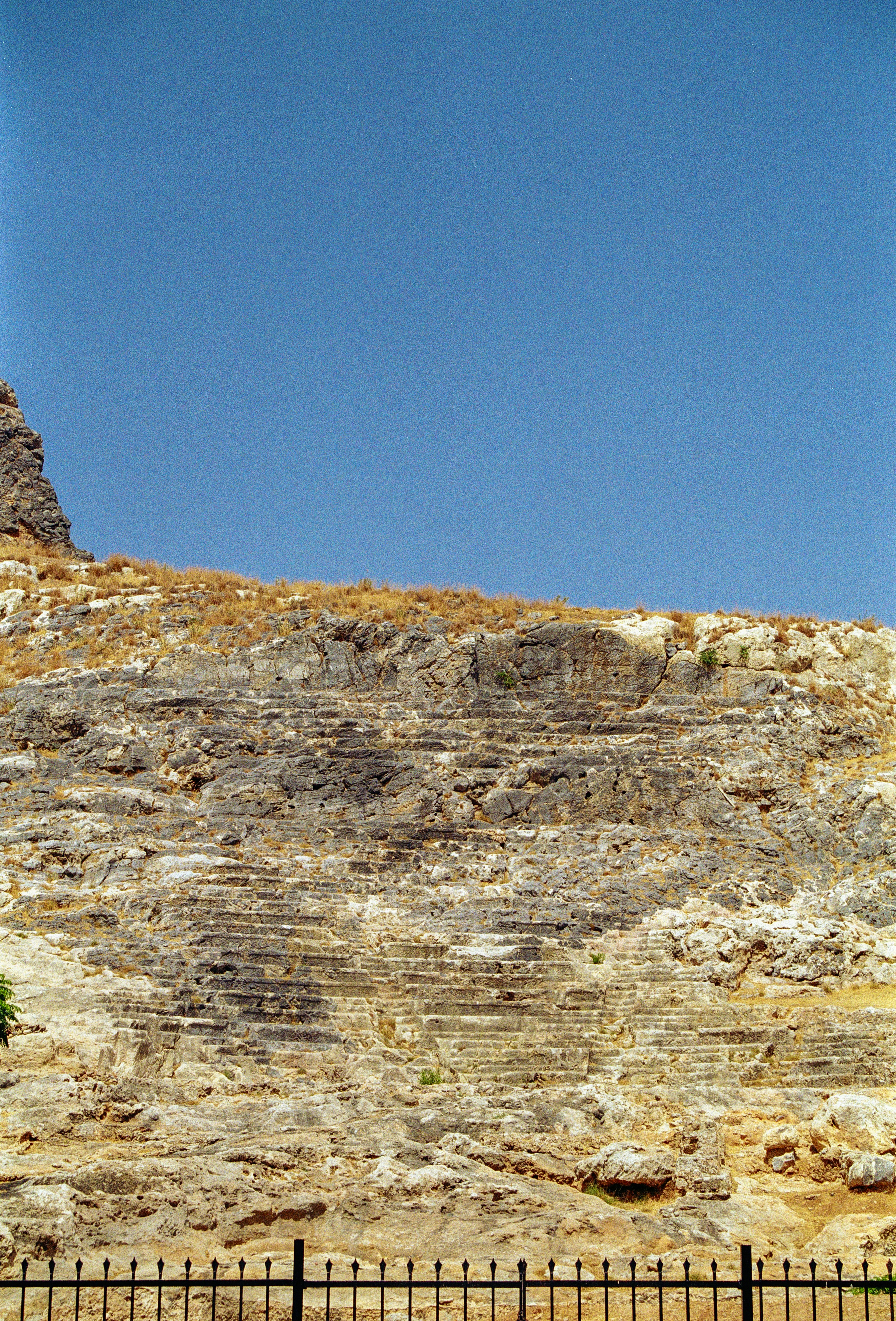 Photograph of a sunlit cliff with distinct horizontal sedimentary layers rising behind a black iron fence under a clear blue sky.