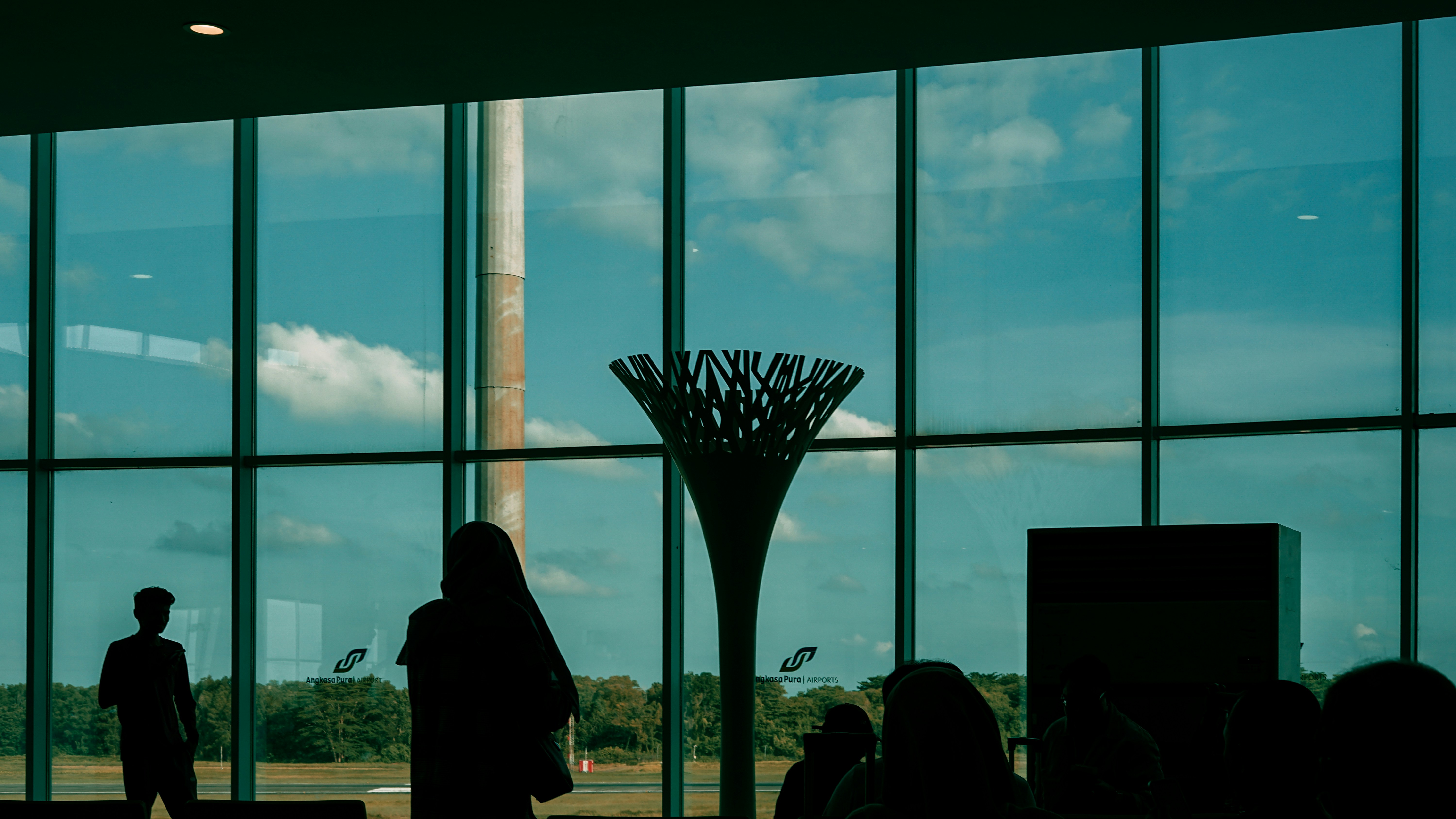 Silhouetted figures stand in a spacious interior, framed by large windows showcasing a vibrant sky and a striking tree-like sculpture.