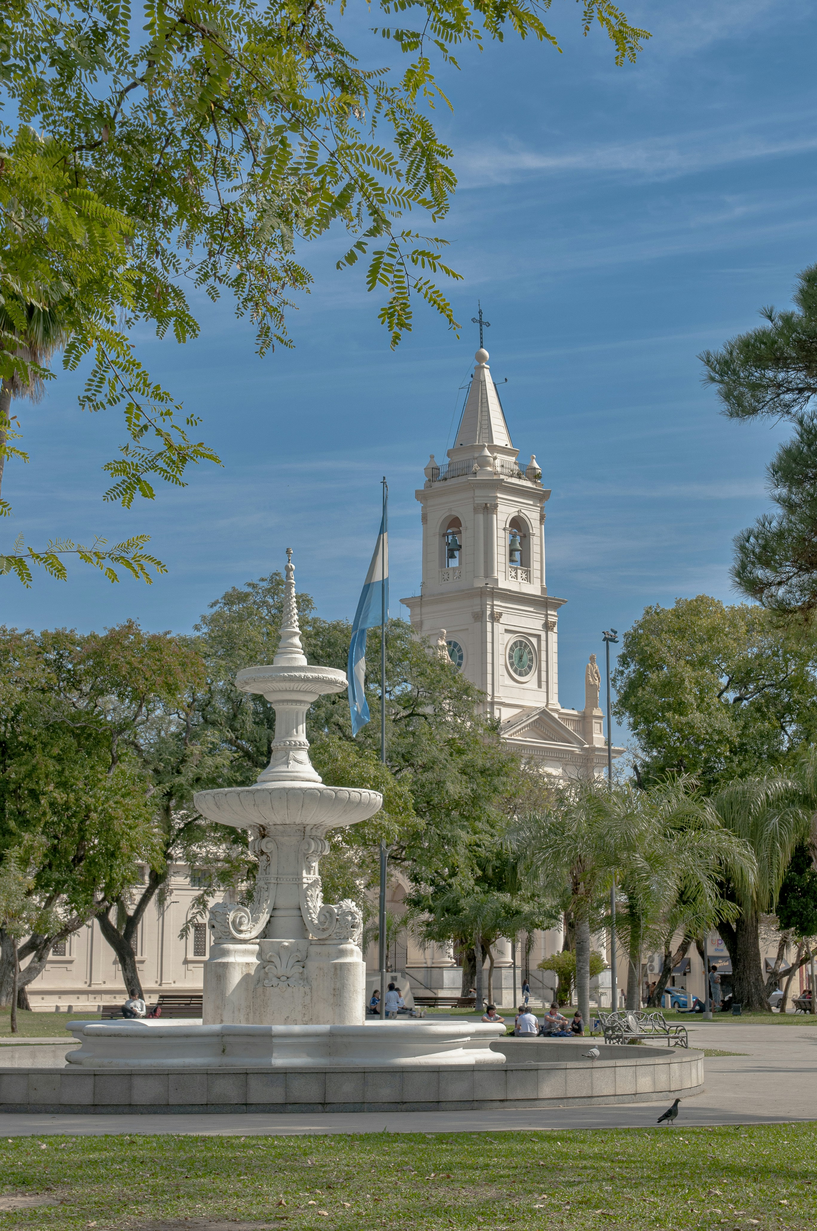 White fountain in a park with a tall church tower under a clear blue sky.