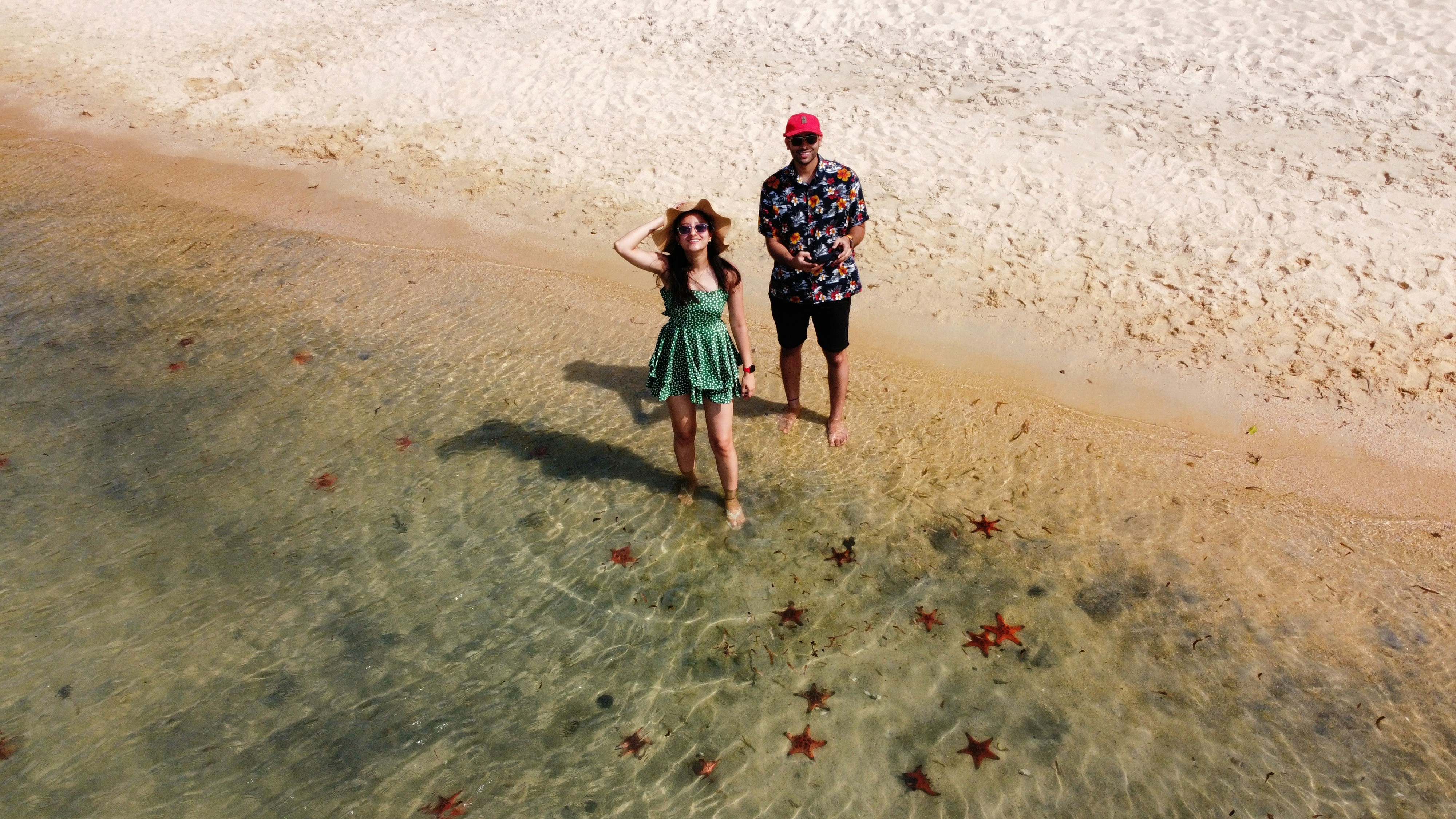 Couple walking through clear shallow water near a sandy beach.