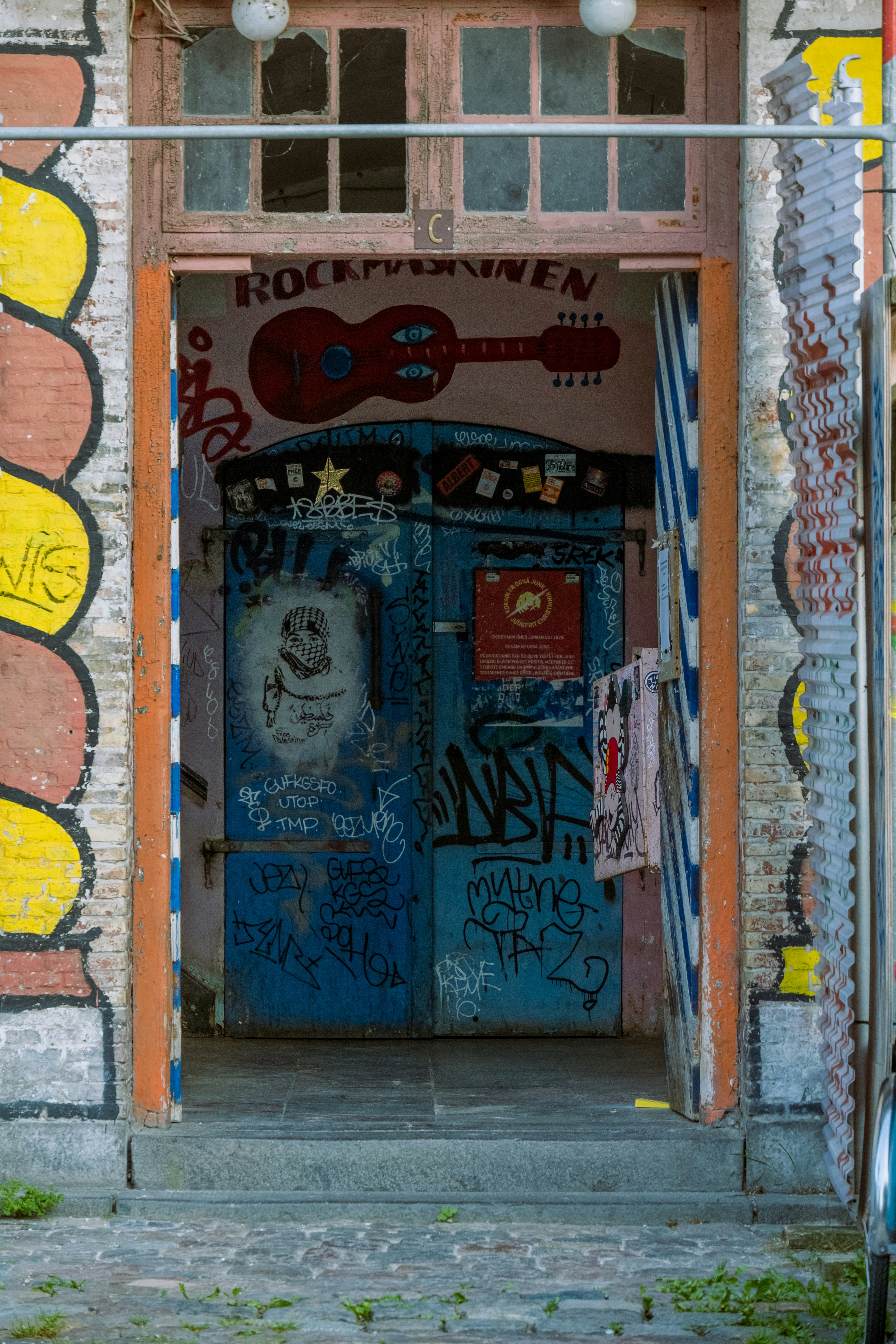 The image shows the entrance of a graffiti-covered building with an urban, artistic vibe. Blue double doors, covered in graffiti, are framed by weathered bricks. Above them, a mural of a red guitar with a face and multiple tuning pegs reads "ROCKMASKINEN," adding a rock-and-roll theme. The surrounding walls feature colorful, abstract shapes, enhancing the eclectic atmosphere.