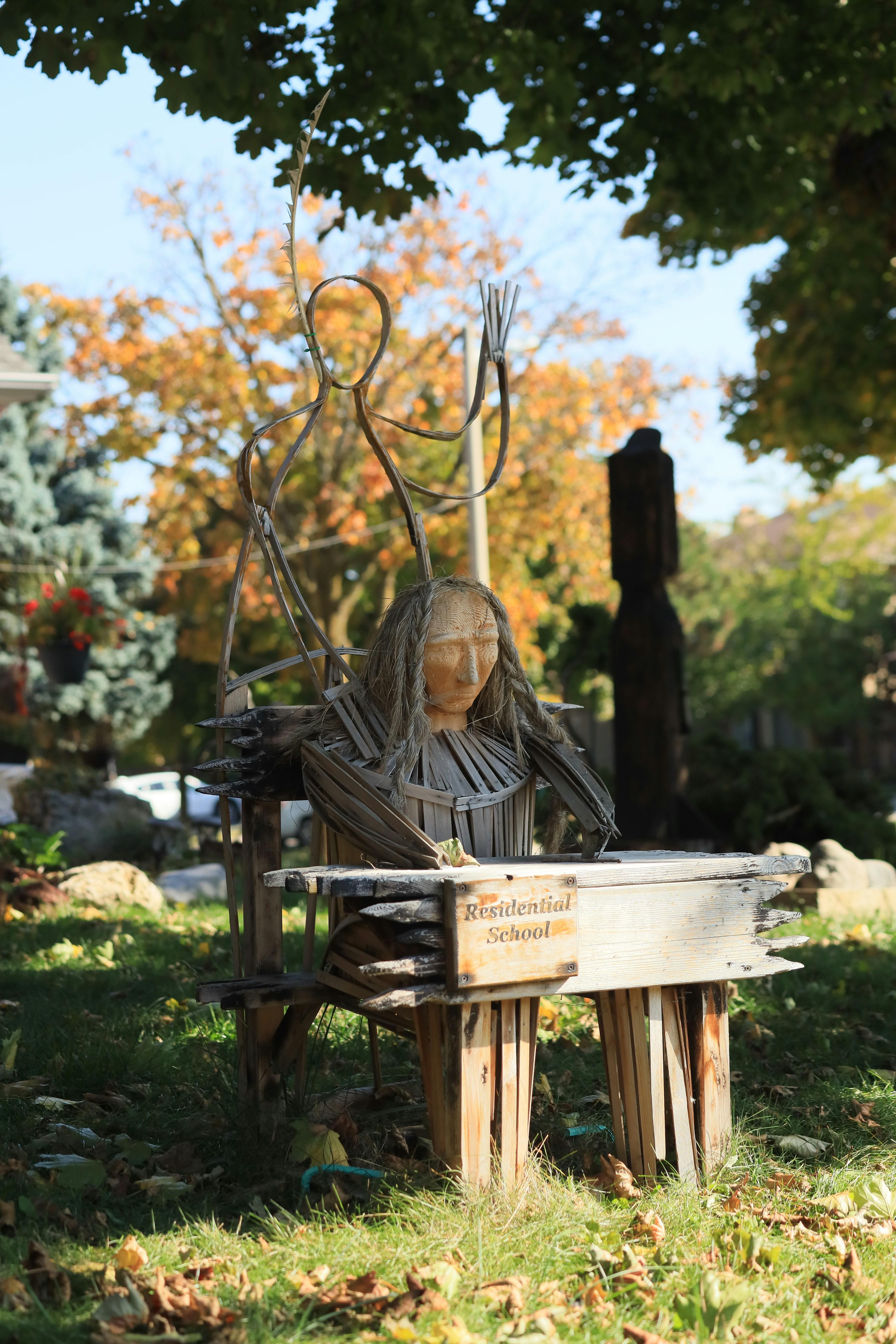 A wooden sculpture depicting a seated figure at a desk, symbolizing the impact of residential schools. The artwork is surrounded by autumn foliage, enhancing its poignant message.