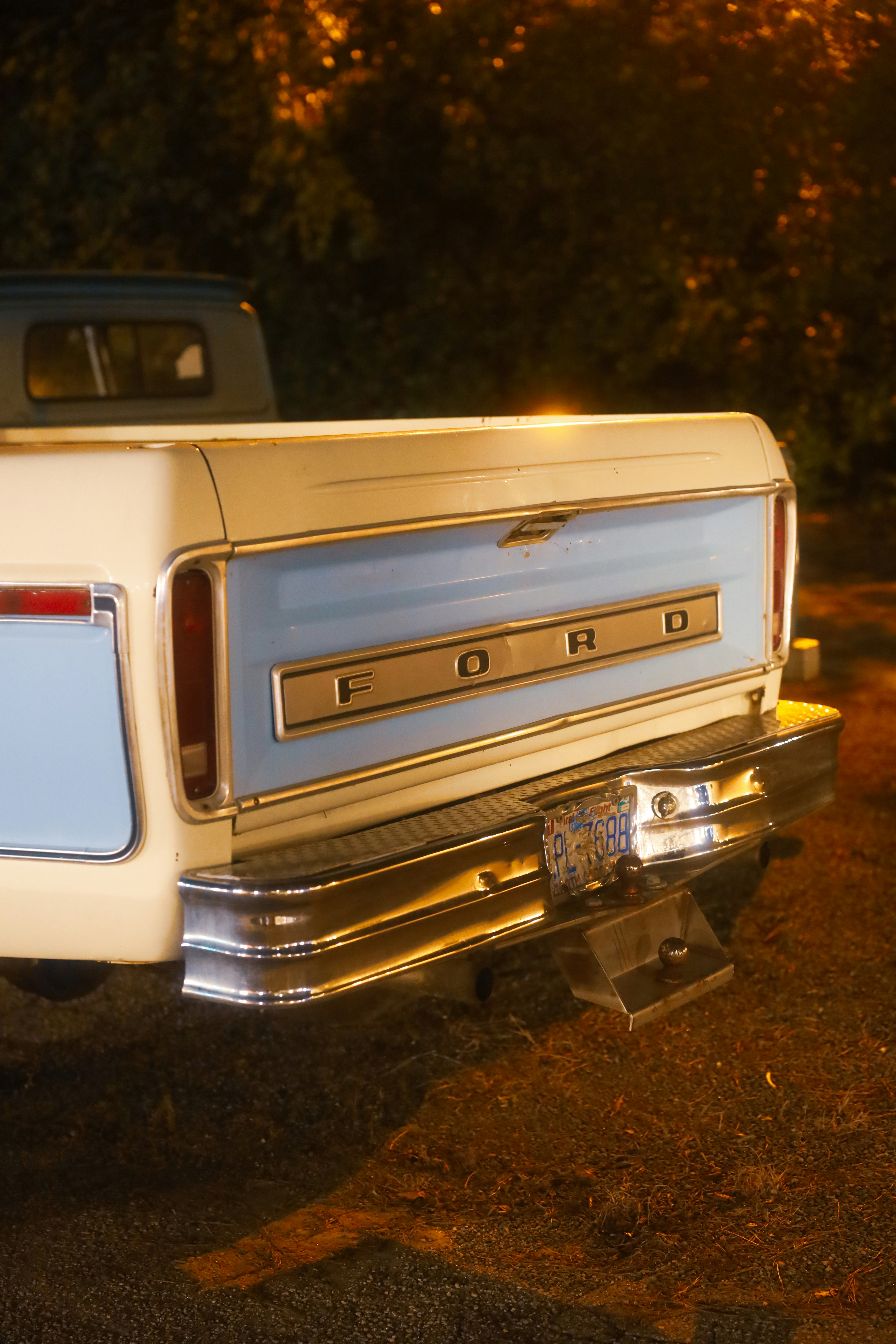 A close-up of a vintage Ford truck tailgate at dusk, chrome bumper gleaming and warm streetlight glow washing over pastel blue paint.