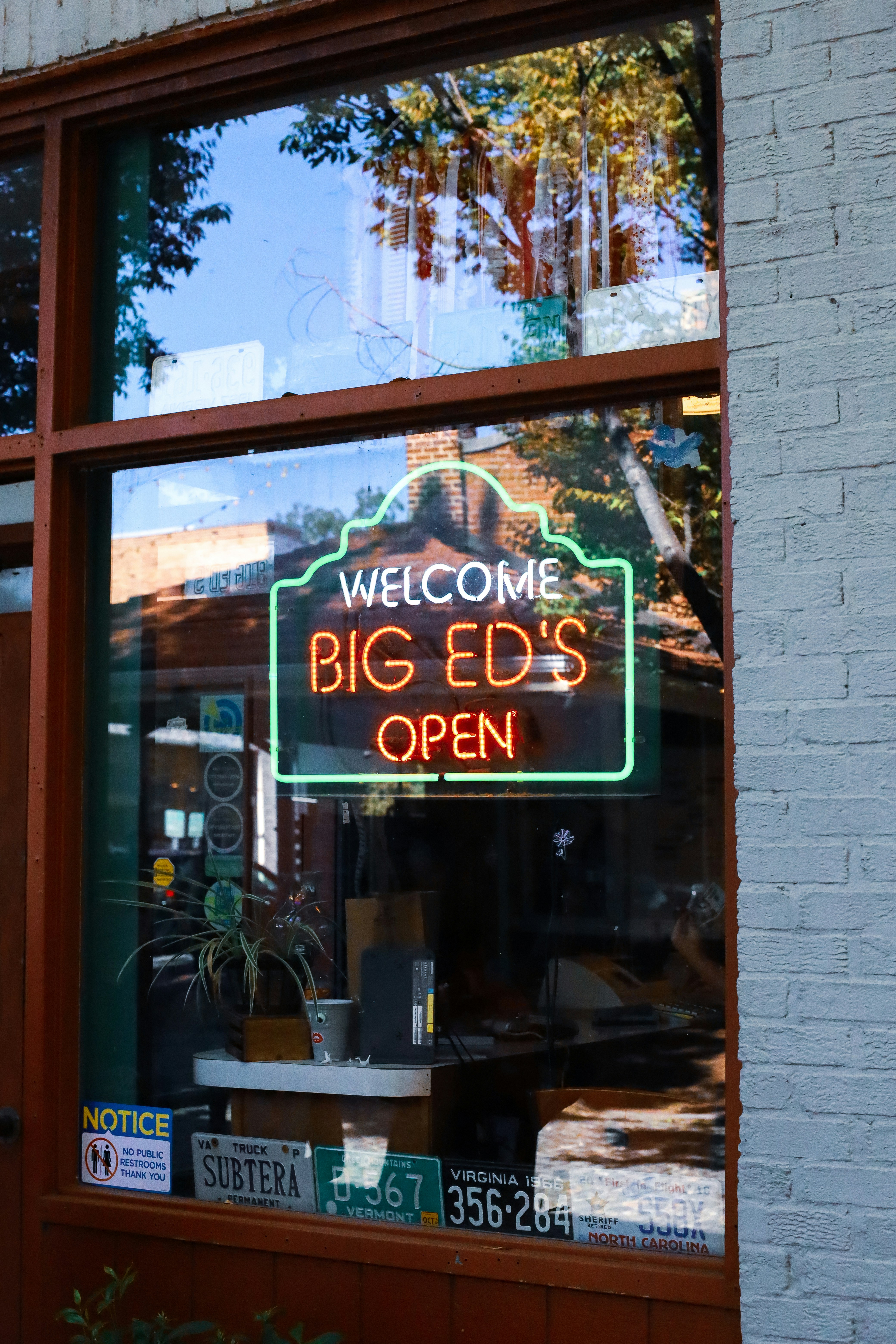 Neon sign reading 'WELCOME BIG ED'S OPEN' illuminates a shop window, framed by rustic wood and greenery. The inviting atmosphere suggests a friendly local business.
