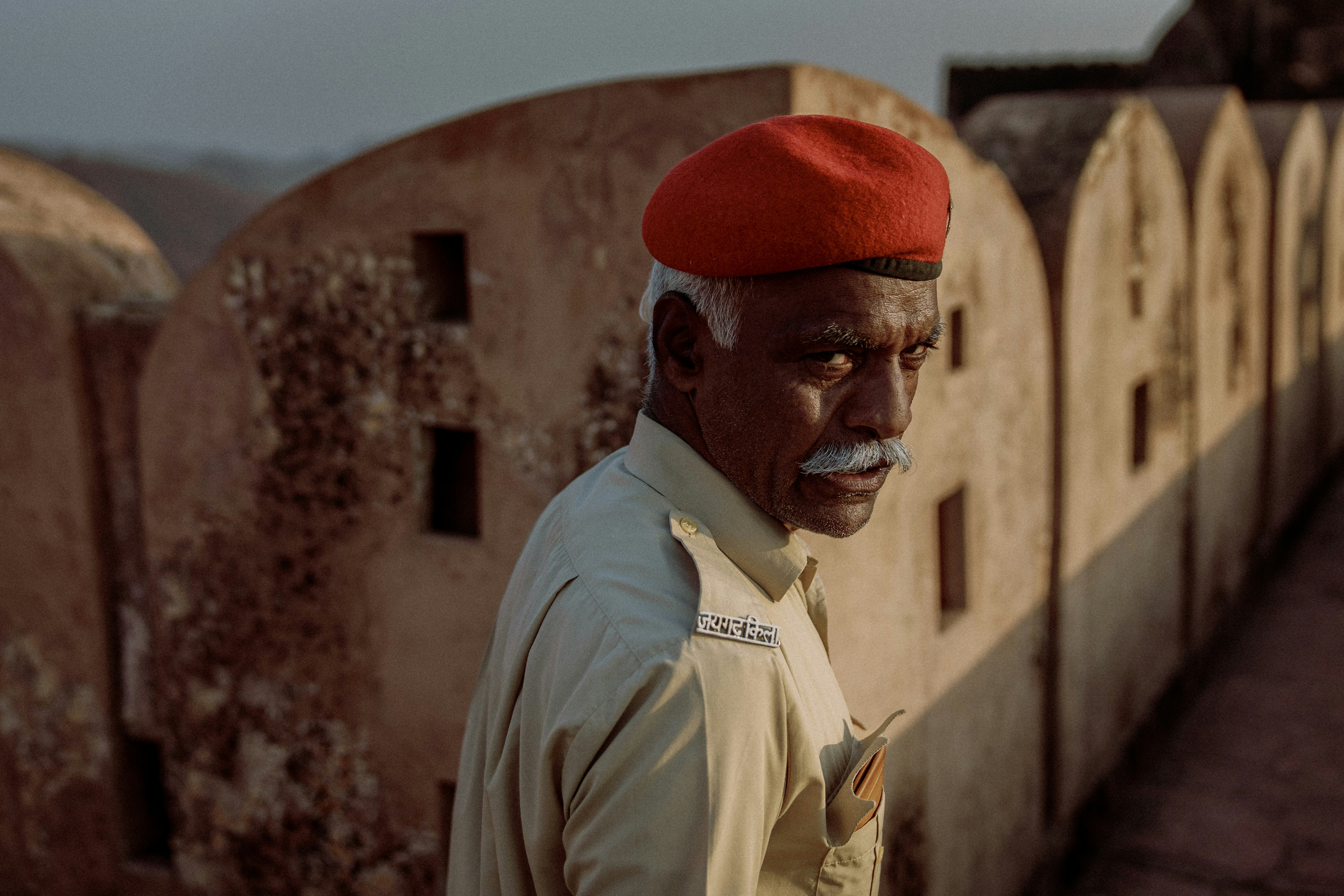 Man in a red hat stands before an ancient, textured wall under soft lighting.