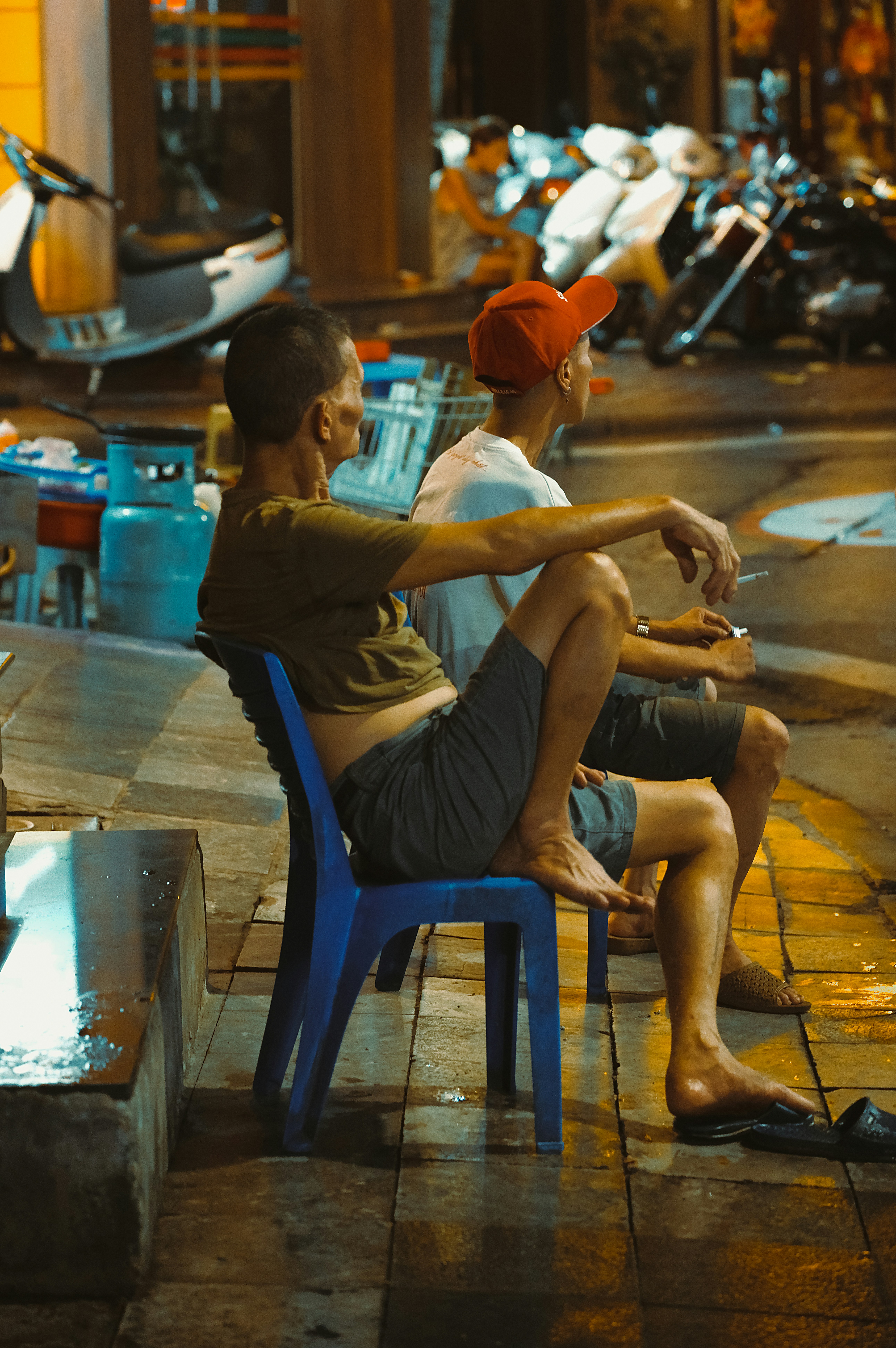 Two men seated casually on plastic chairs, engaged in conversation on a bustling street at night. The scene captures the essence of urban life.