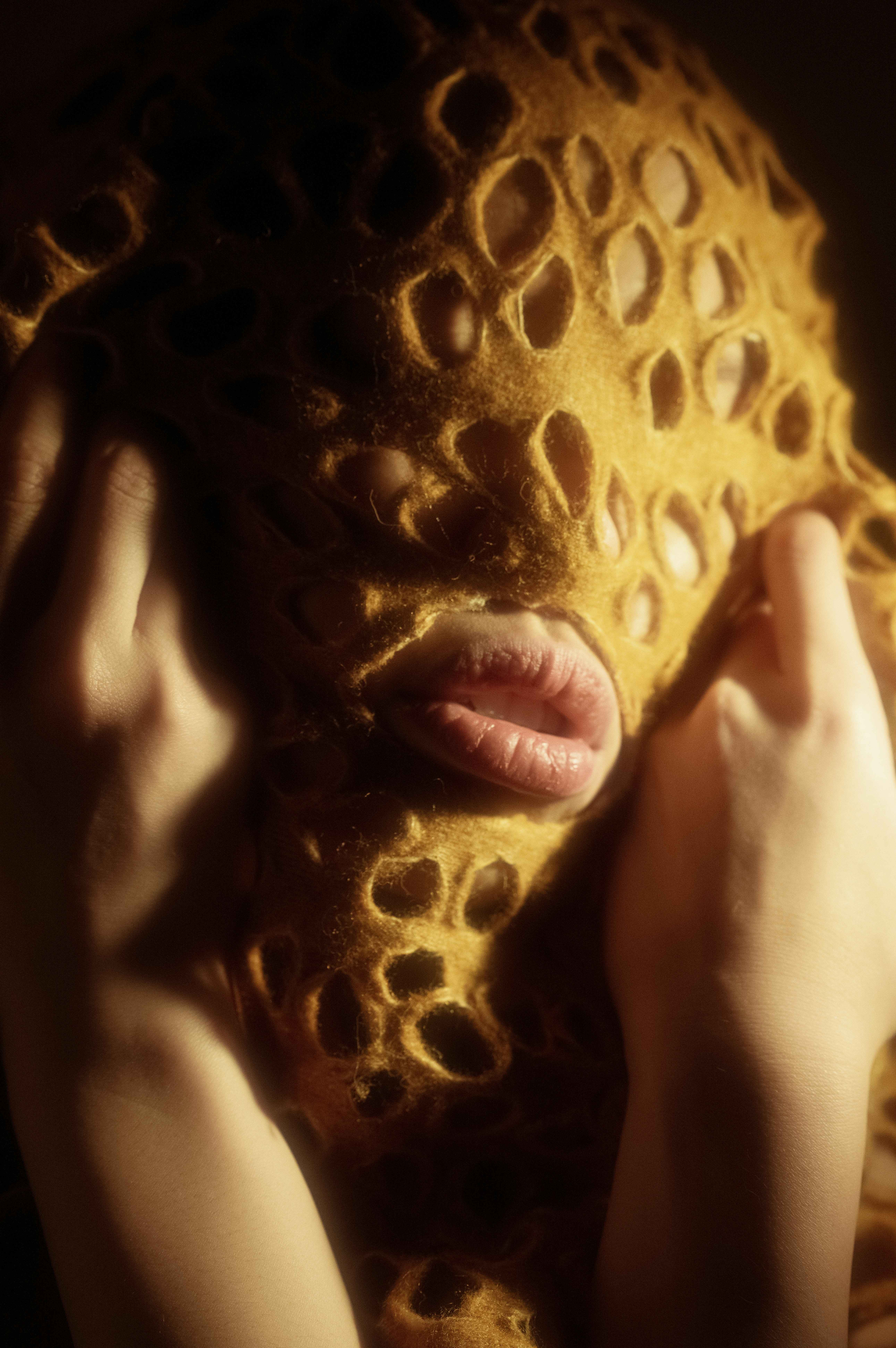 Close-up of a leopard-patterned fabric mask with circular openings, lips visible through the holes and hands framing the scene. Warm, directional light highlights texture and contrast.