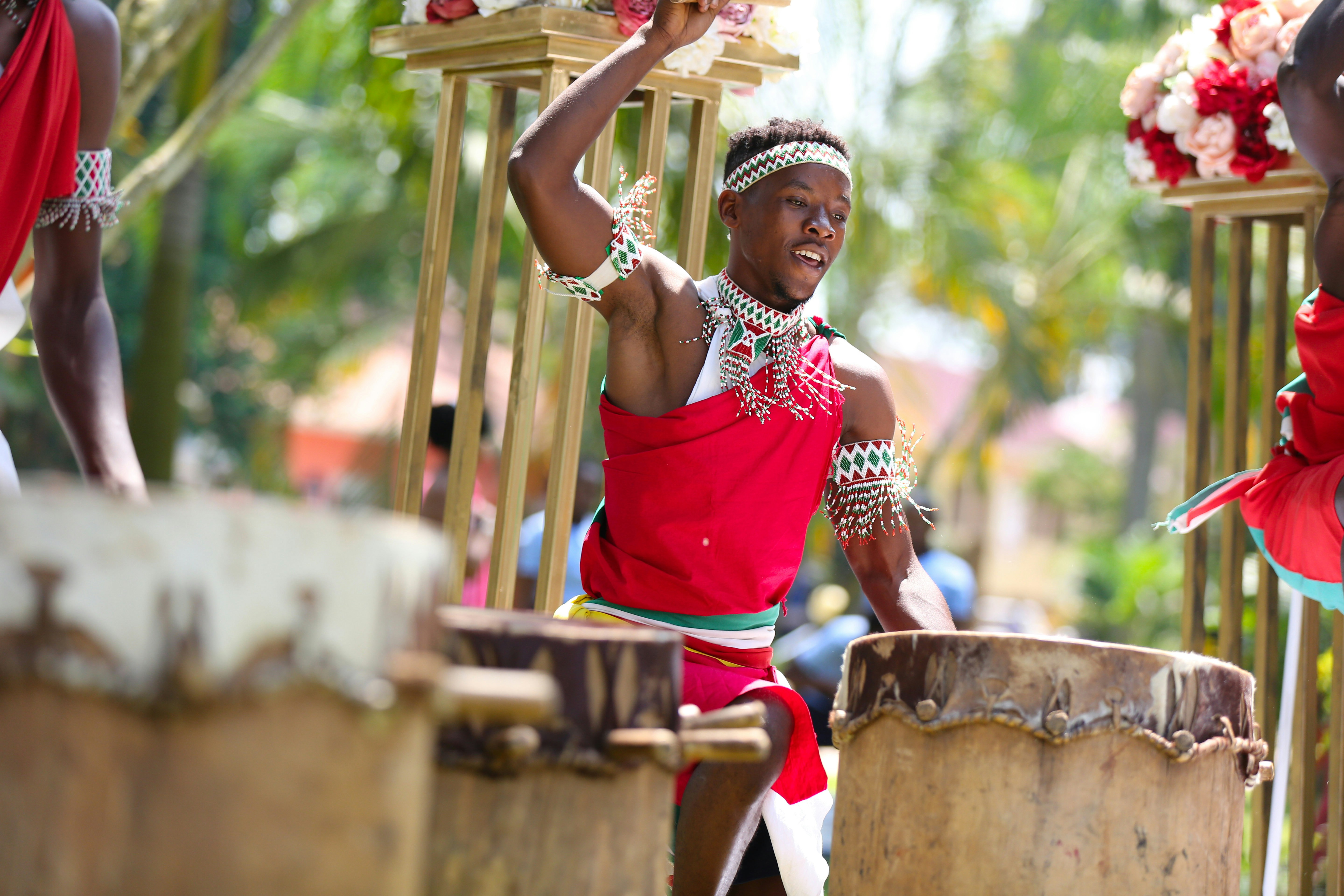 A drummer in vibrant attire passionately performs on traditional drums at an outdoor Ugandan wedding.