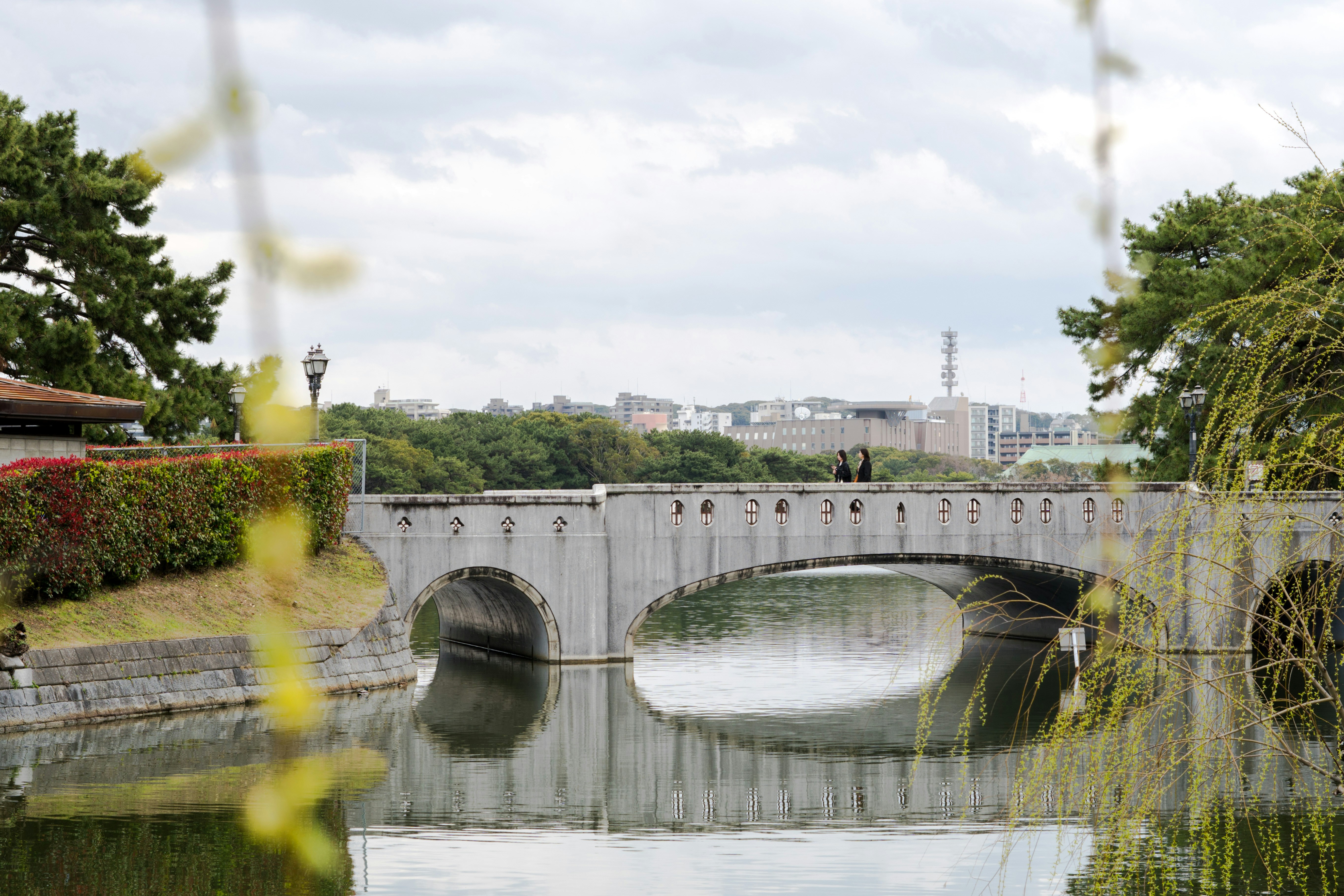 A stone bridge arches gracefully over a reflective body of water, framed by lush greenery and urban silhouettes in the background.